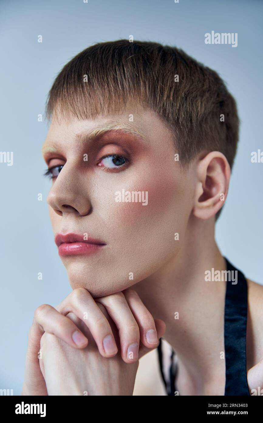 queer model with makeup looking at camera on grey backdrop, androgynous ...