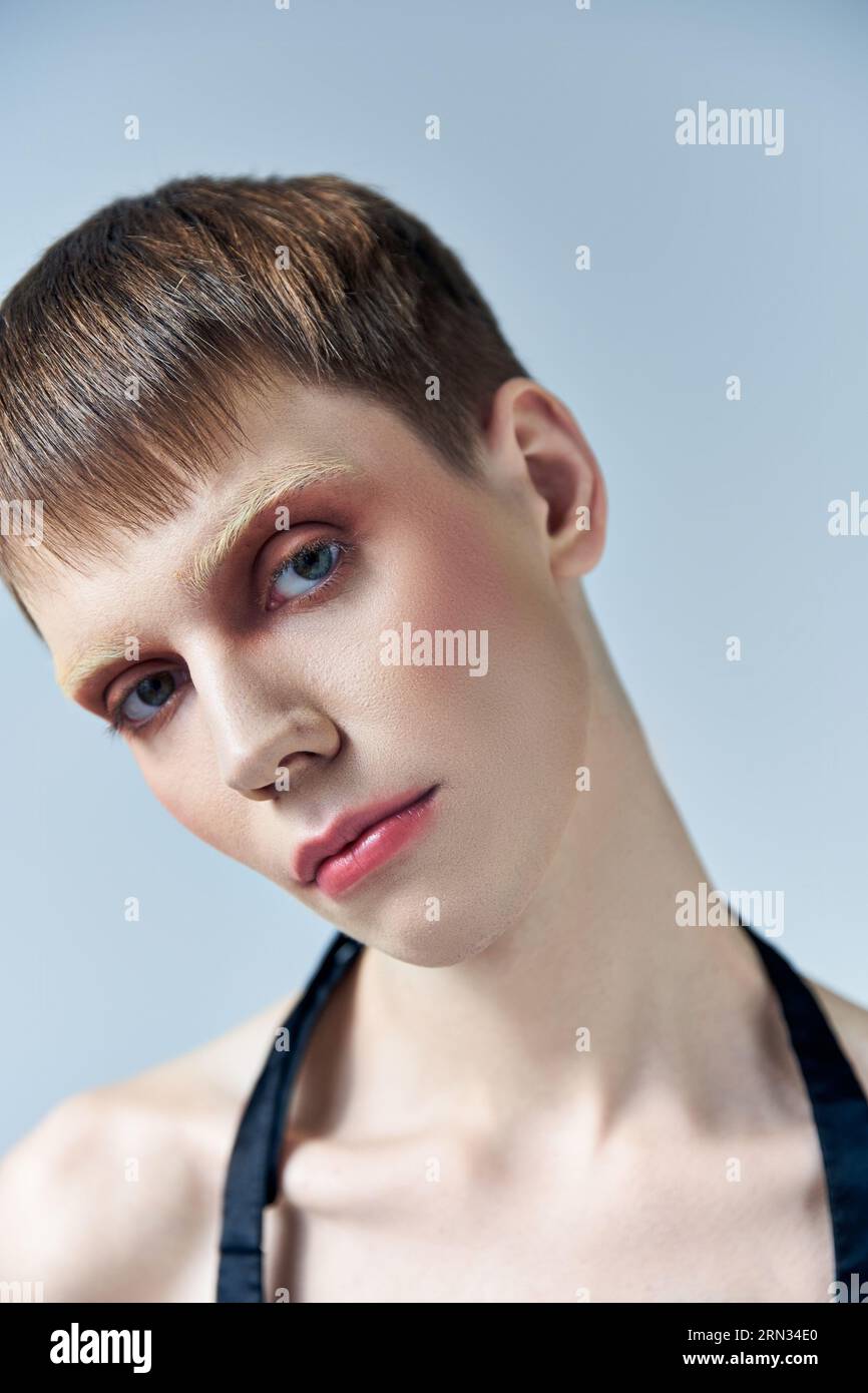 portrait, androgynous person with makeup looking at camera on grey, beauty and visage, queer ...