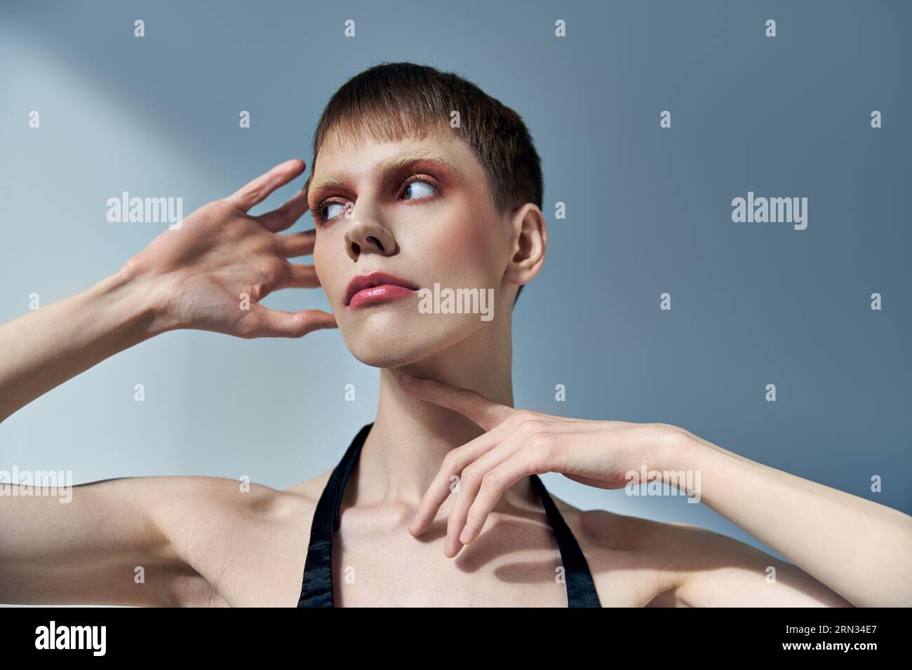 queer person with makeup looking away and posing on grey backdrop ...