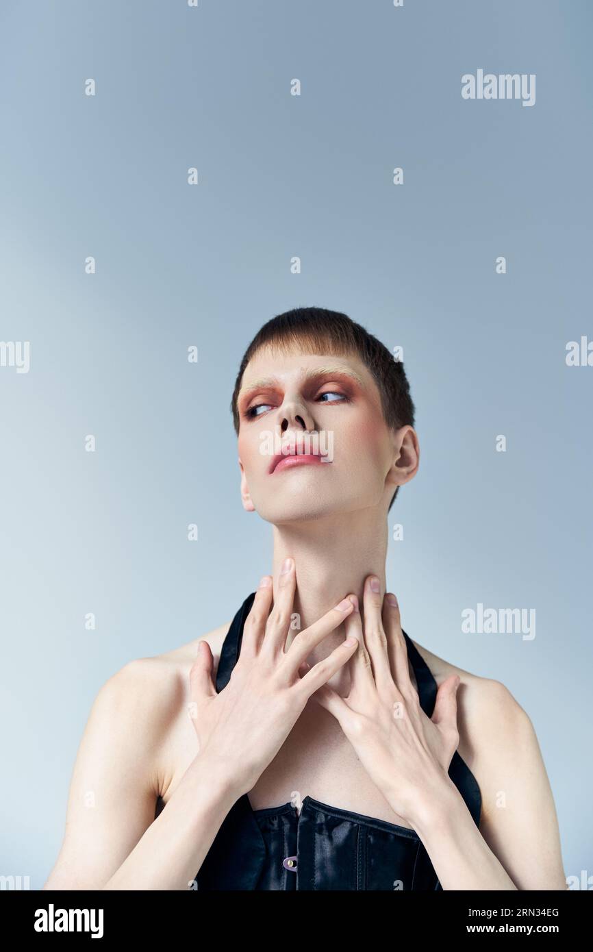 portrait, queer model with makeup touching neck and posing on grey ...