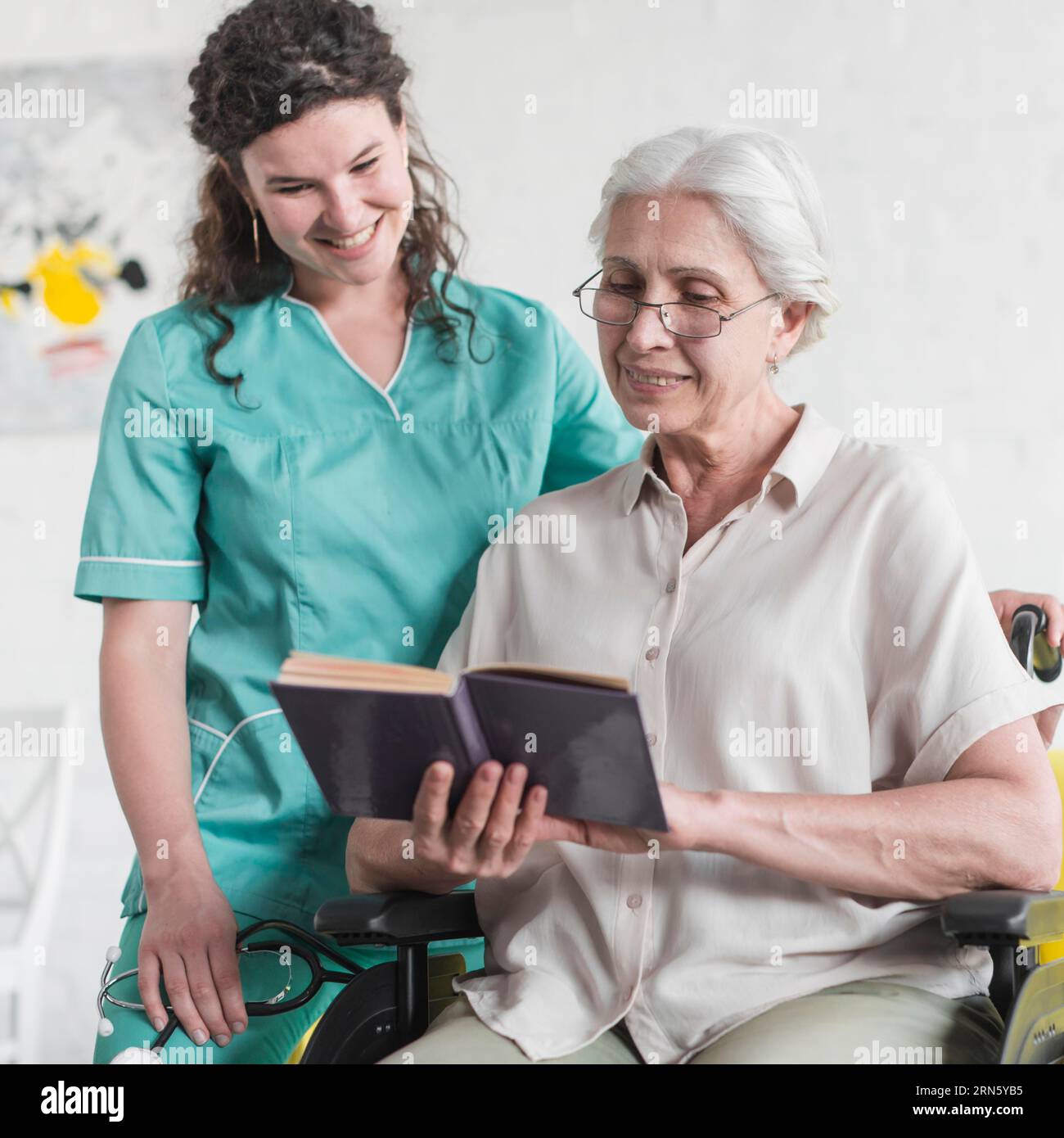 Nurse standing disabled senior woman reading book Stock Photo - Alamy