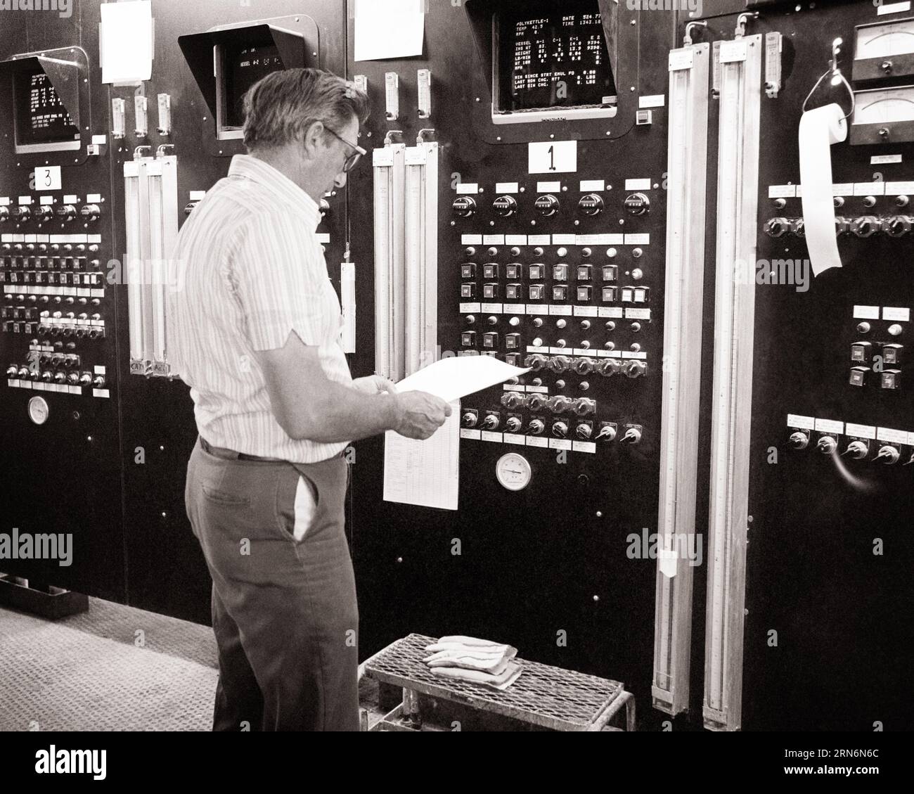 1970s MAN READING A REPORT STANDING BEFORE A COMPLEX CONTROL PANEL IN A ...