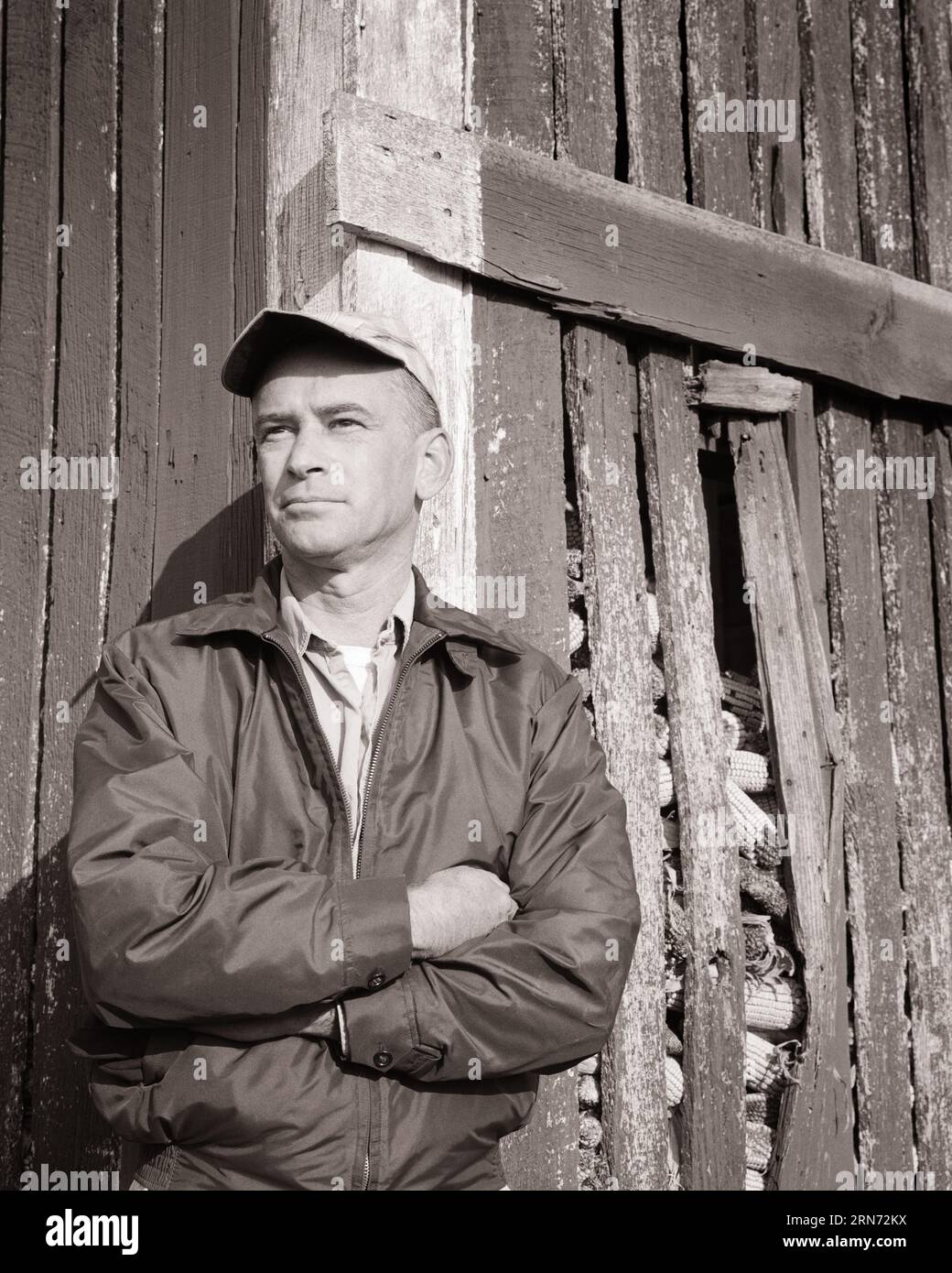 1960s MAN FARMER STANDING BY CORN CRIB WEARING JACKET CAP ARMS CROSSED ...