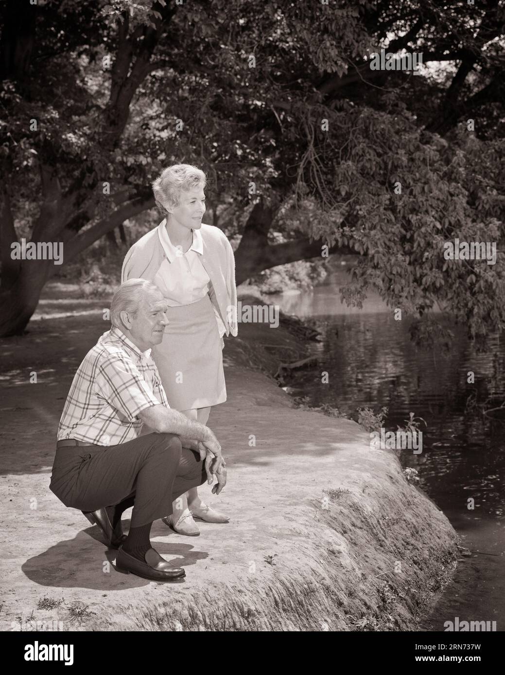 1960s SENIOR COUPLE IN PARK MAN KNEELING BESIDE POND WOMAN STANDING ...
