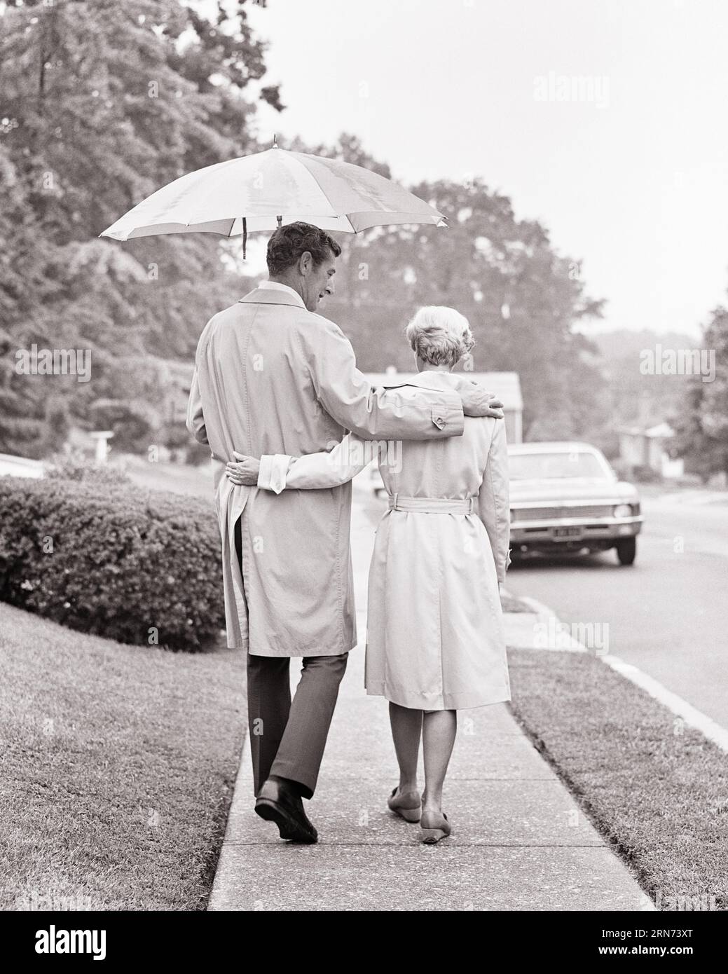 1970s MATURE COUPLE WALKING ON SUBURBAN SIDEWALK ARM IN ARM BACK VIEW ...