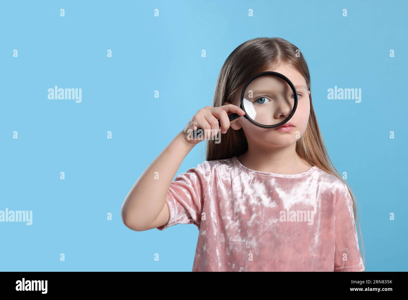 Cute little girl looking through magnifier glass on light blue background. Space for text Stock ...