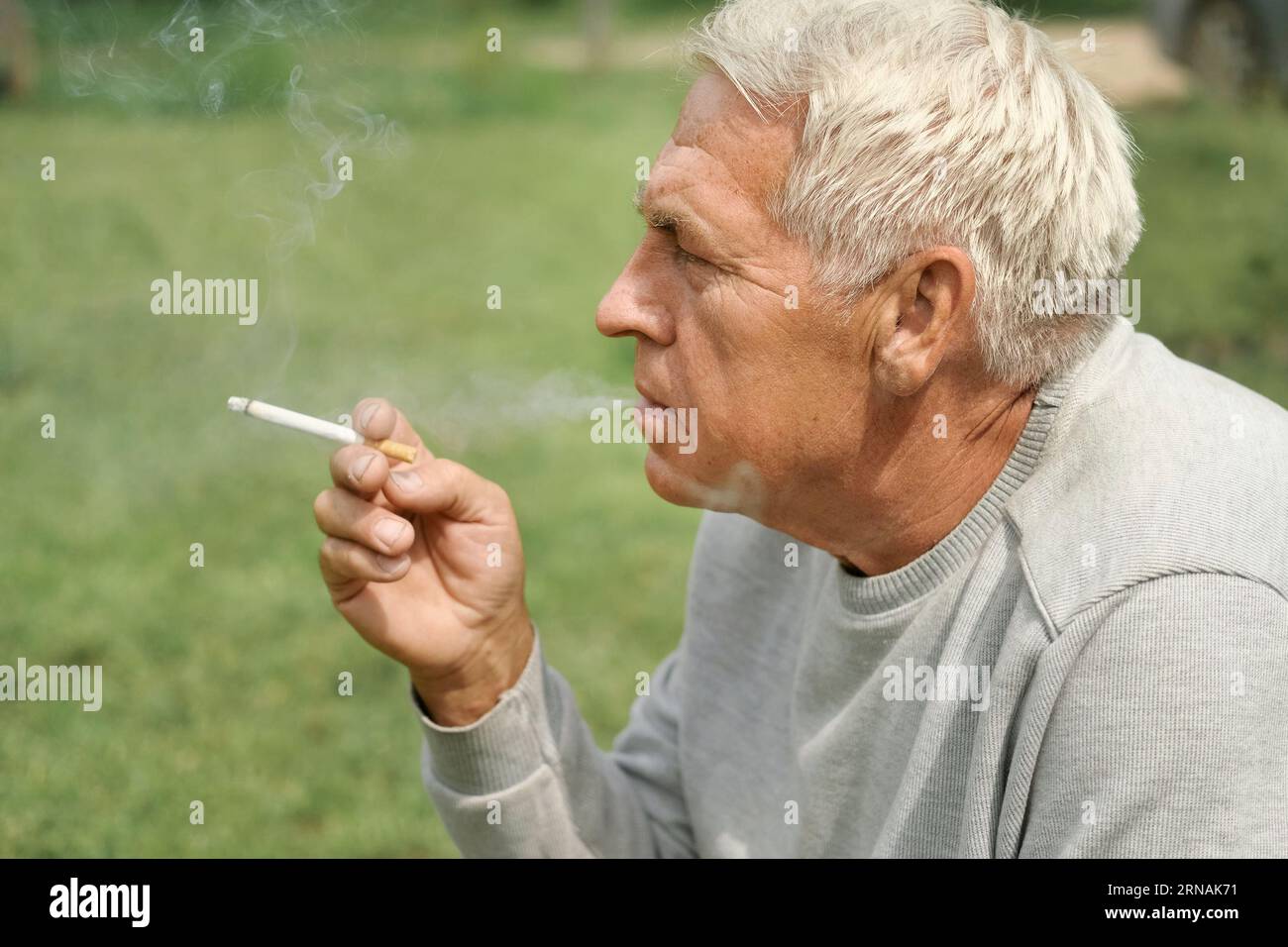 Senior man lighting up a cigarette outdoors against green nature ...