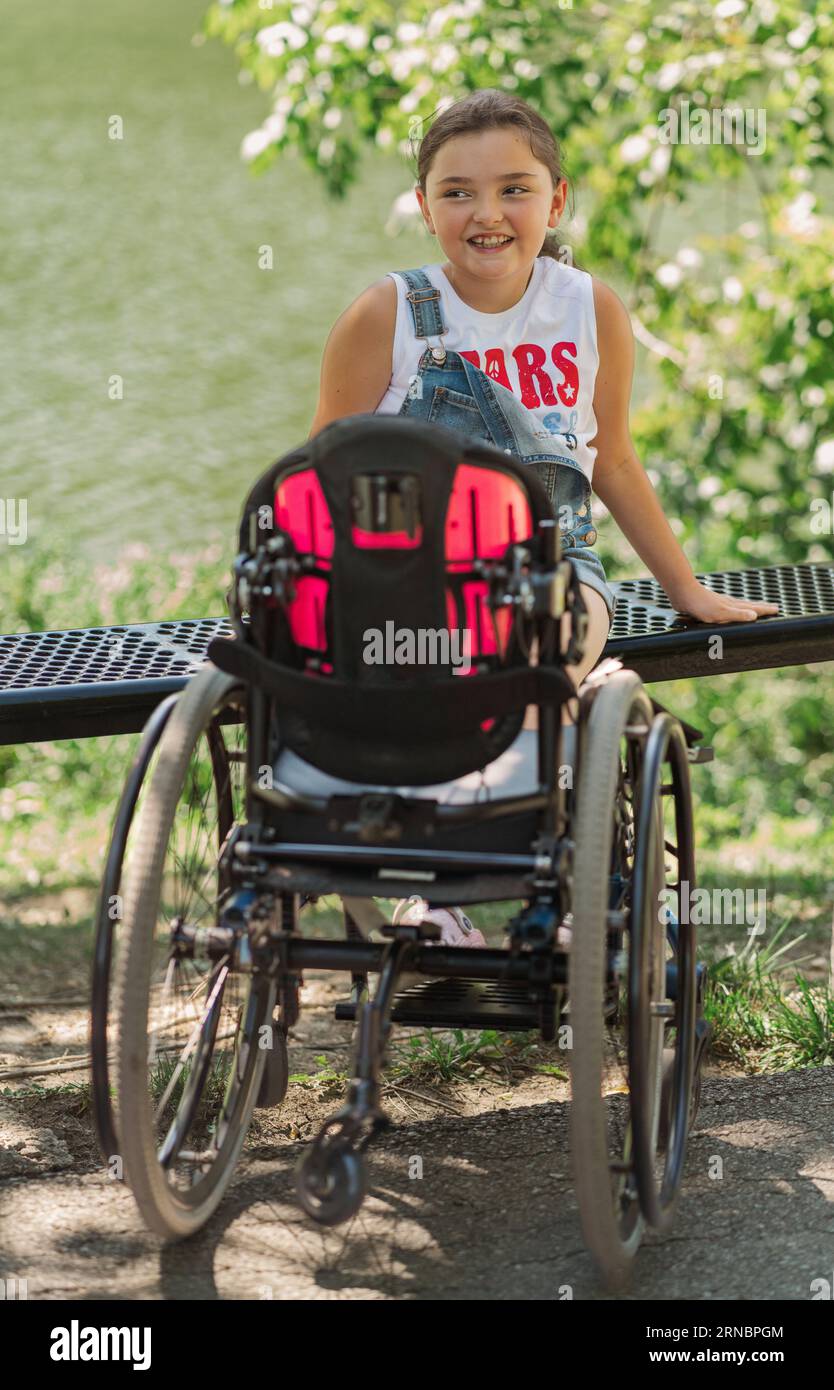 A girl sitting on a bench by the lake with her wheelchair in front ...