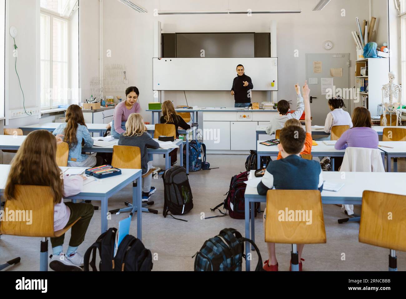 Male and female teacher teaching students sitting on bench in classroom ...