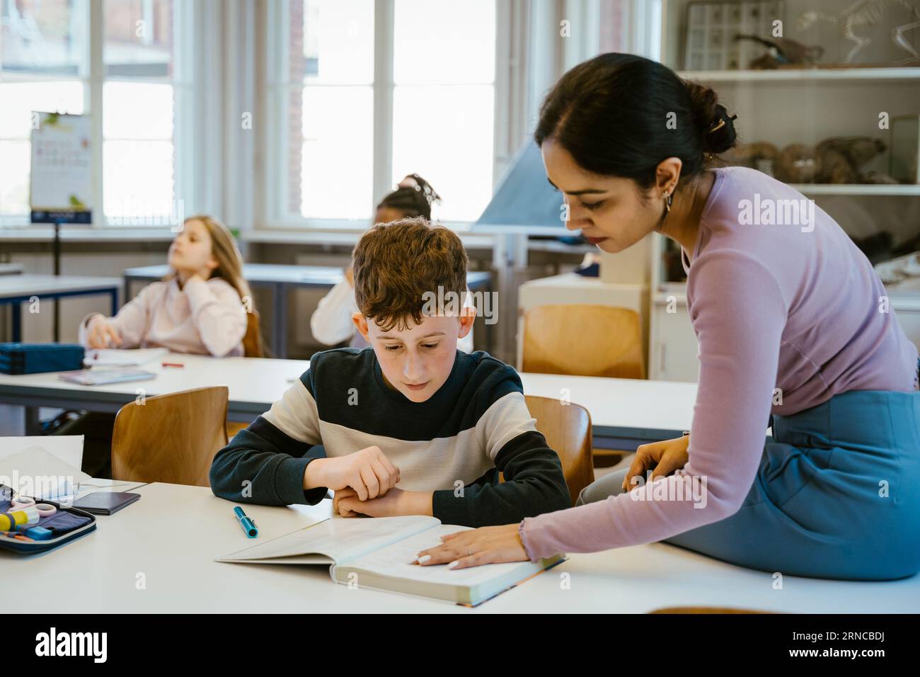 Female teacher helping schoolboy reading in book while sitting on desk ...