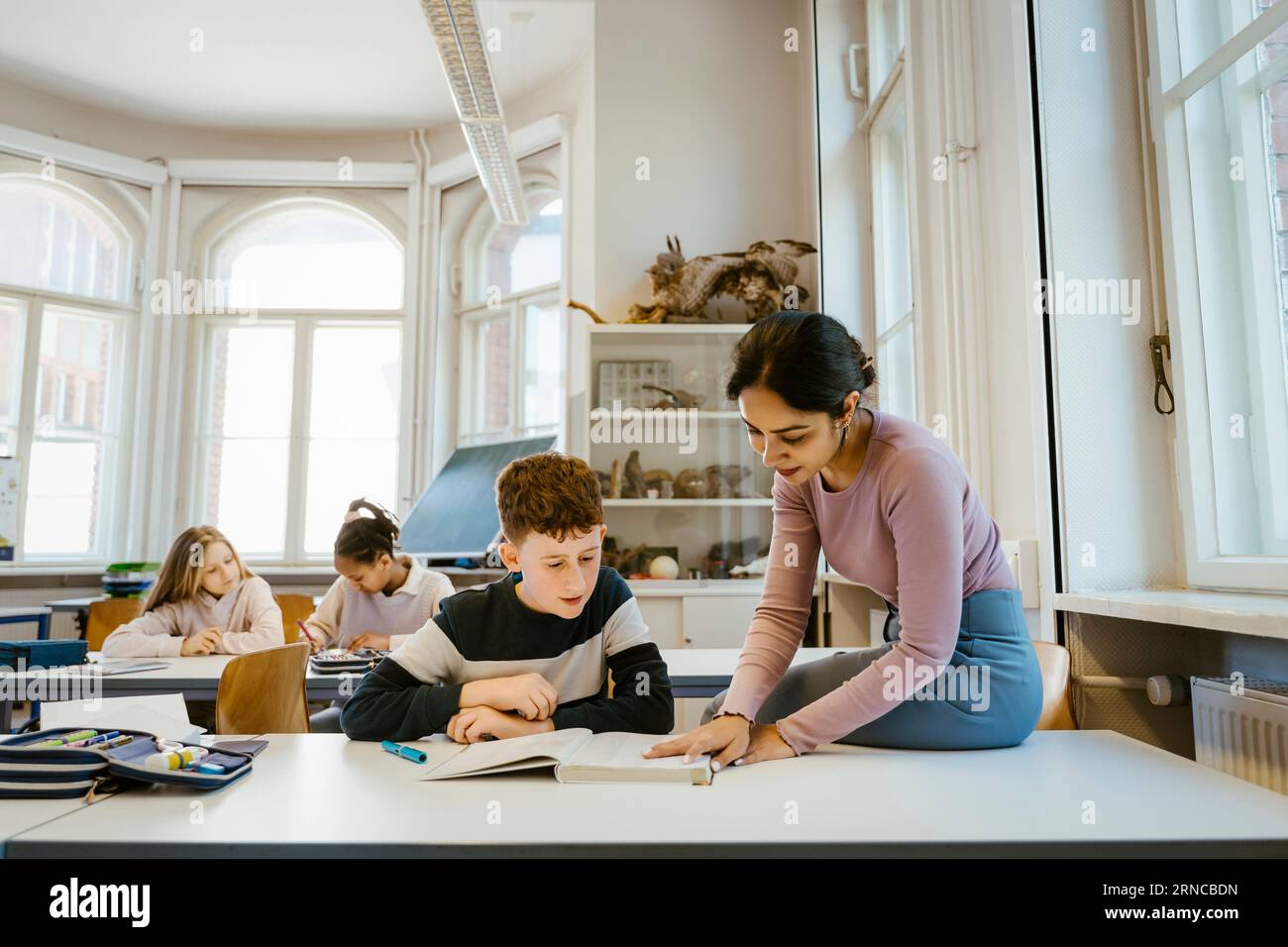 Female teacher assisting schoolboy reading book while sitting at desk ...