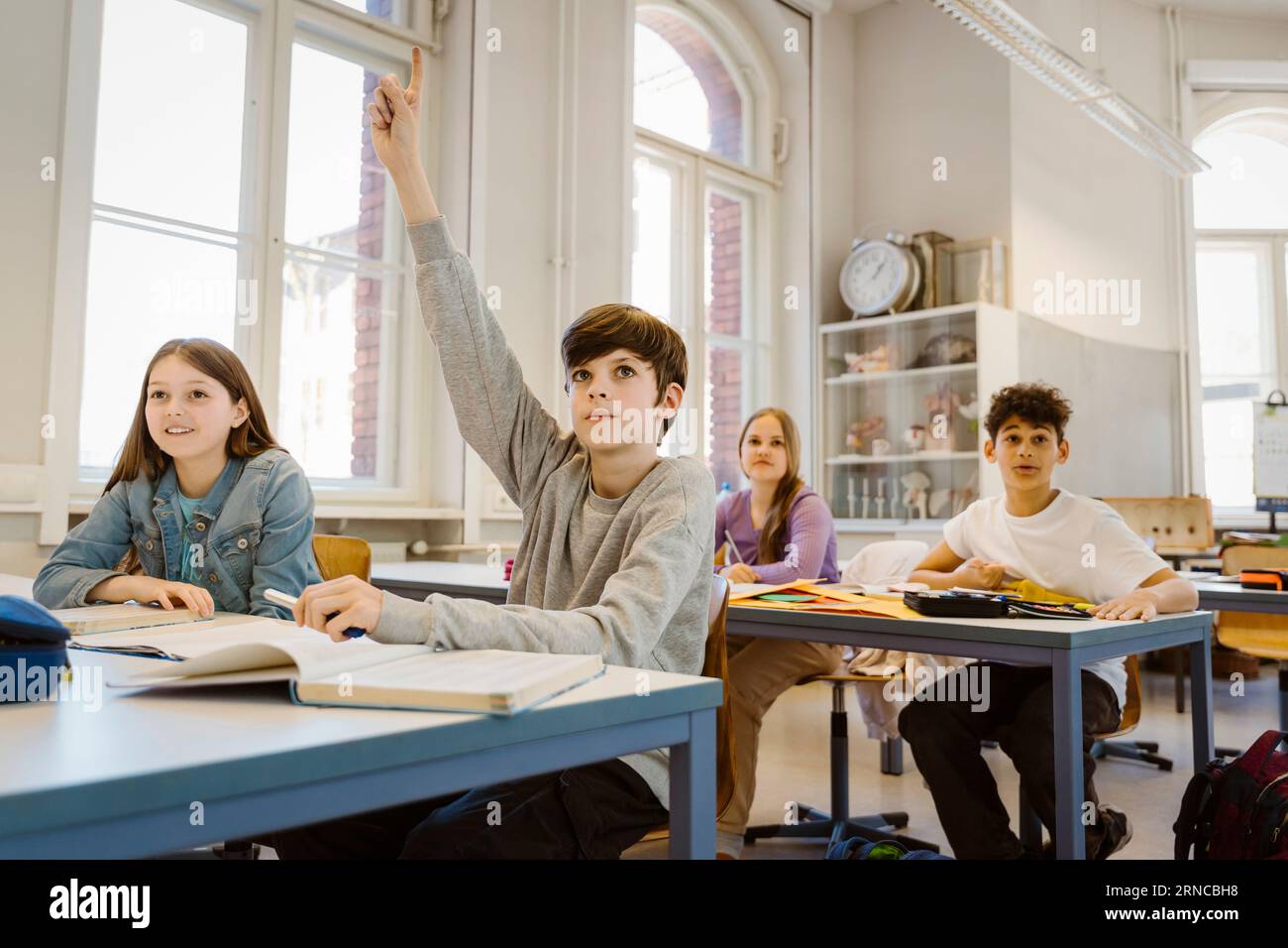 Boy raising hand while sitting with female friend at desk in classroom ...