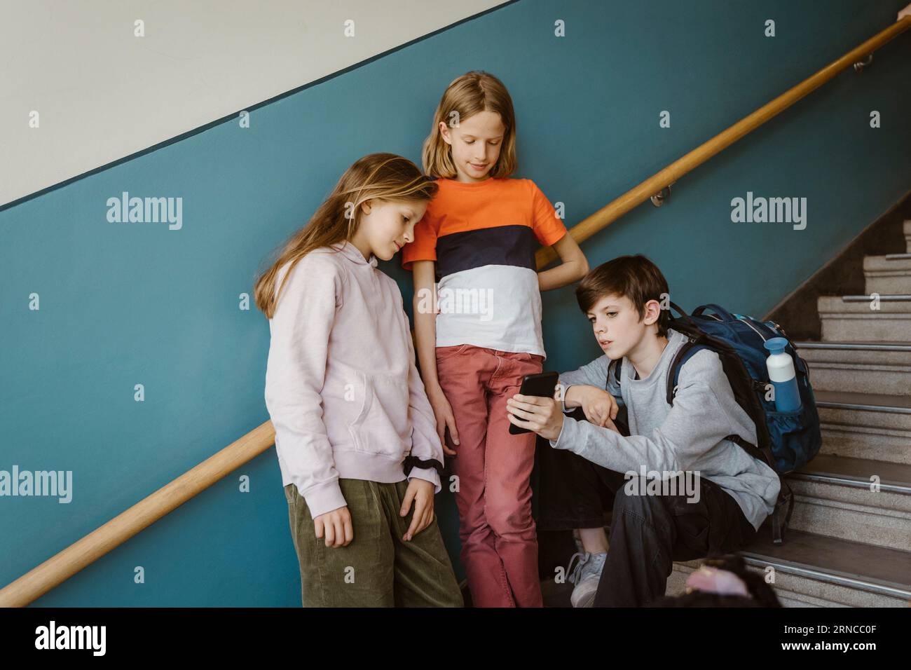 Boy sharing smart phone with female friend standing against wall on ...