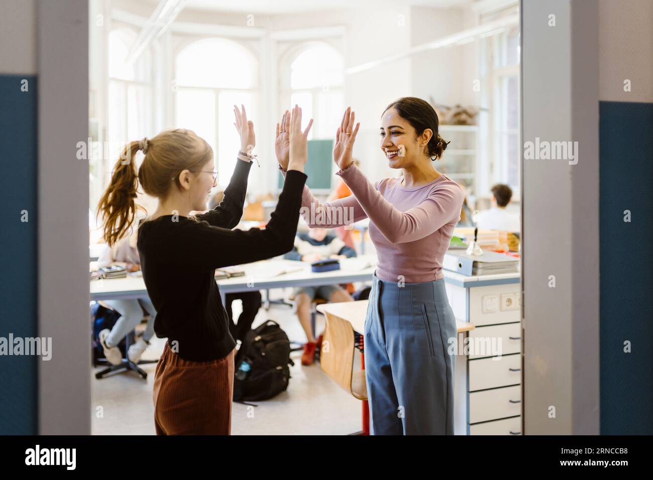 Smiling female teacher giving high-five to girl standing in classroom ...