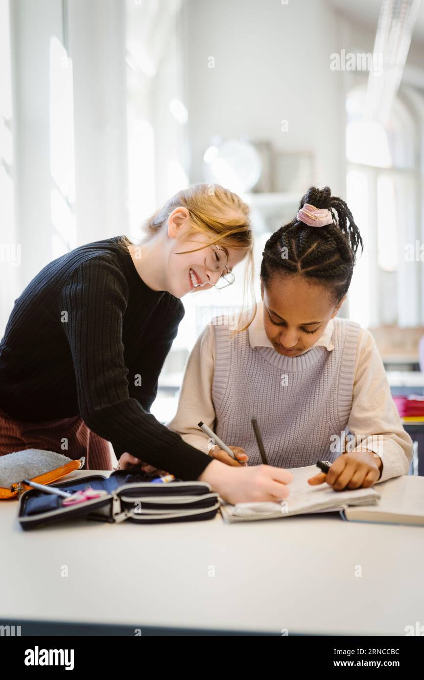 Smiling girl with female friend writing in book while studying in ...