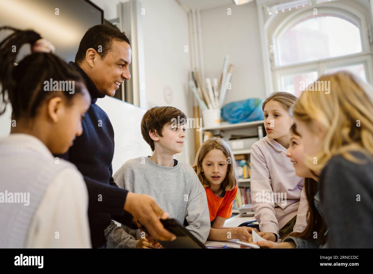 Male teacher sharing tablet PC with students discussing in classroom ...