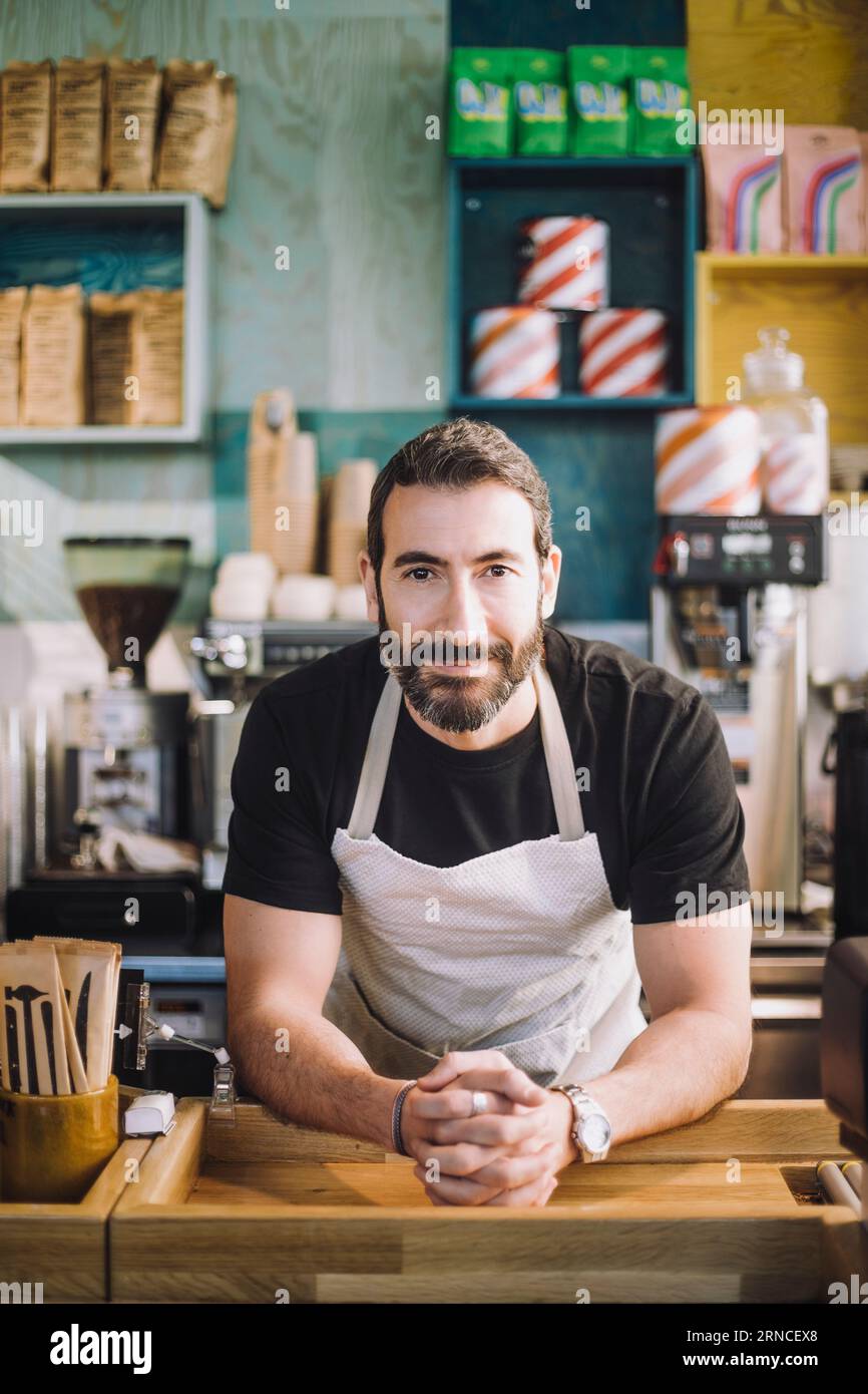 Portrait of confident male retail clerk leaning on checkout counter at ...