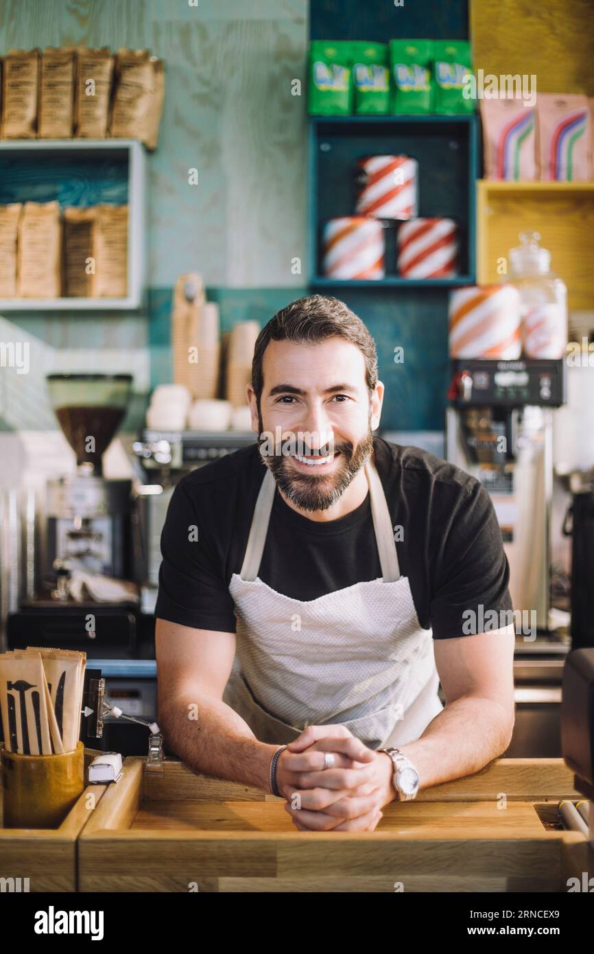 Portrait of smiling male retail clerk with hands clasped leaning on ...