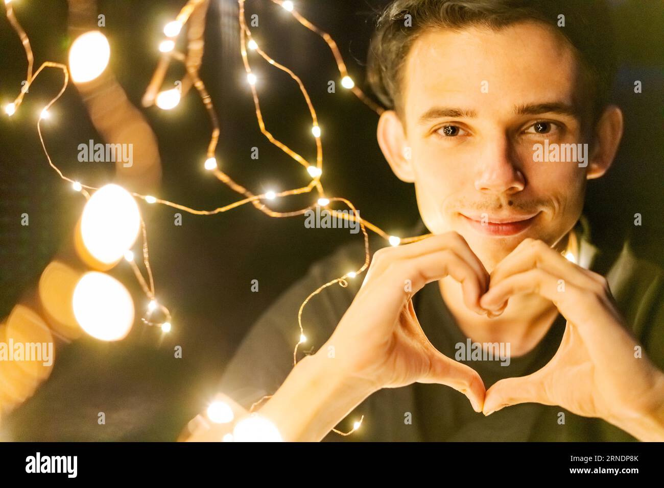 a young guy at night holding a glowing garland shows a heart gesture ...