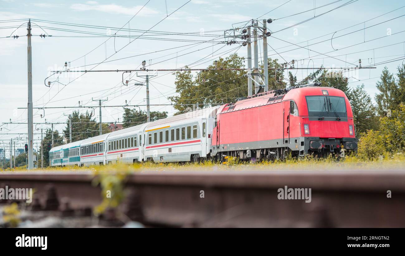 Passenger train with modern red locomotive and four coaches is rushing through the station of ...