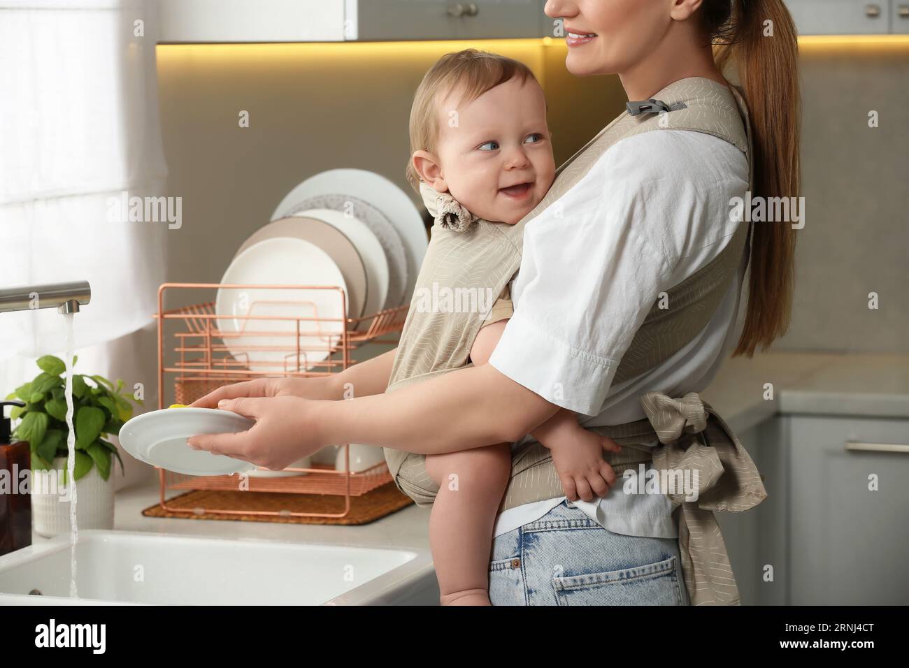 Mother holding her child in sling (baby carrier) while washing plates ...