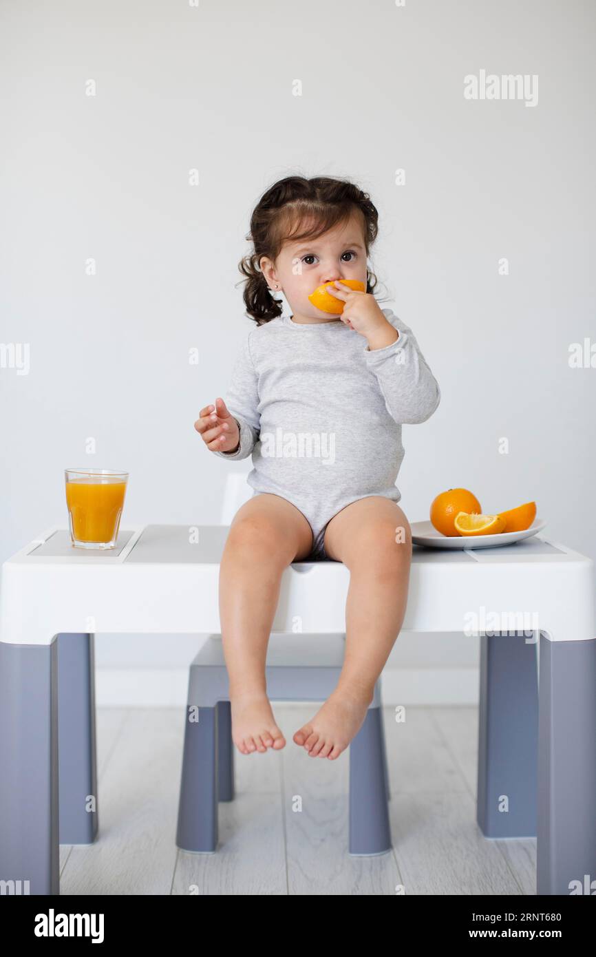 Sitting table baby girl eating orange Stock Photo - Alamy