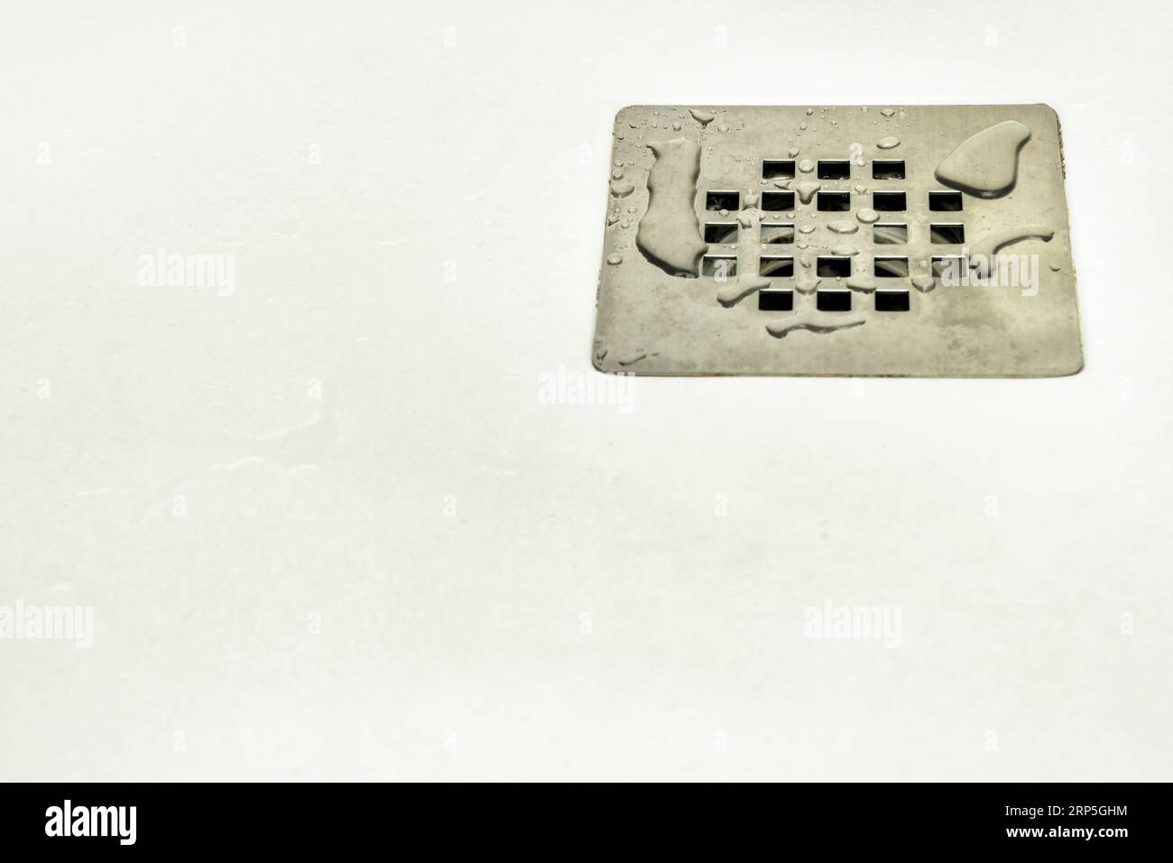 A square metal shower drain with water drops on a white shower tray ...