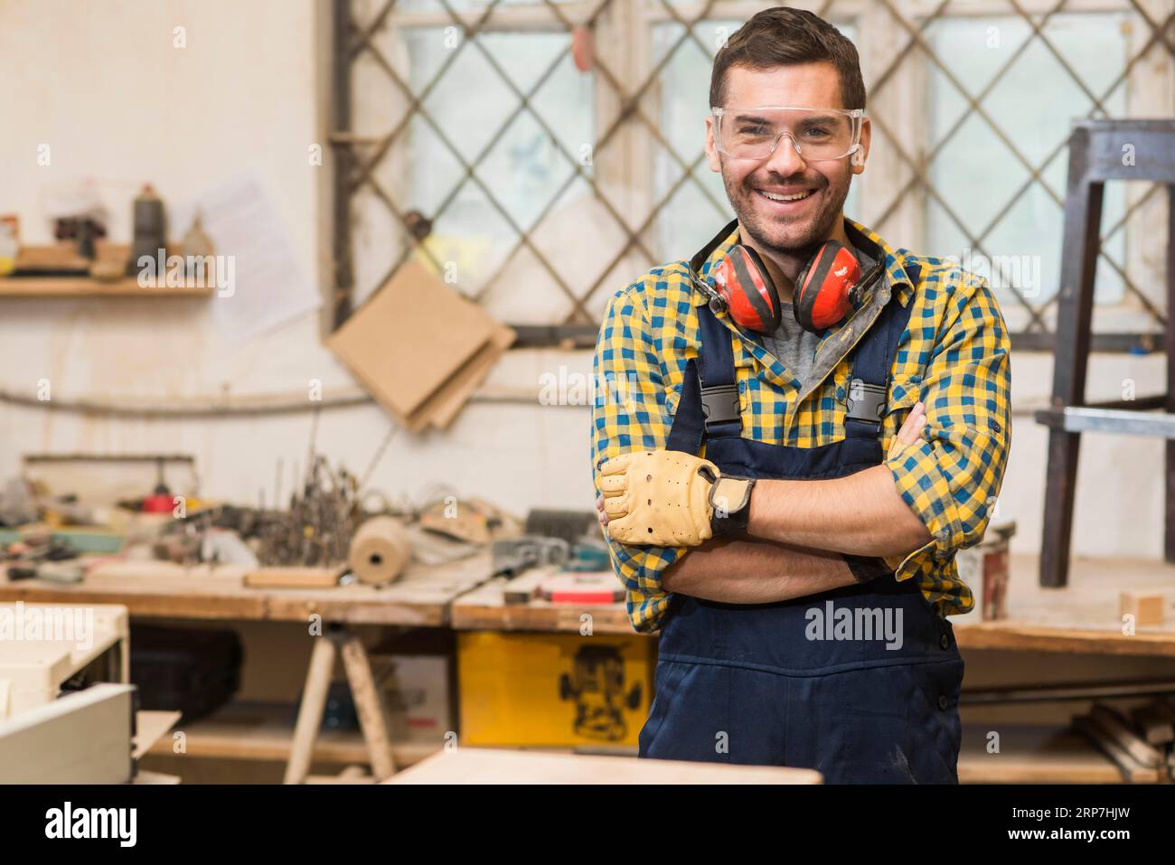 Smiling male carpenter wearing safety glasses standing front workbench ...