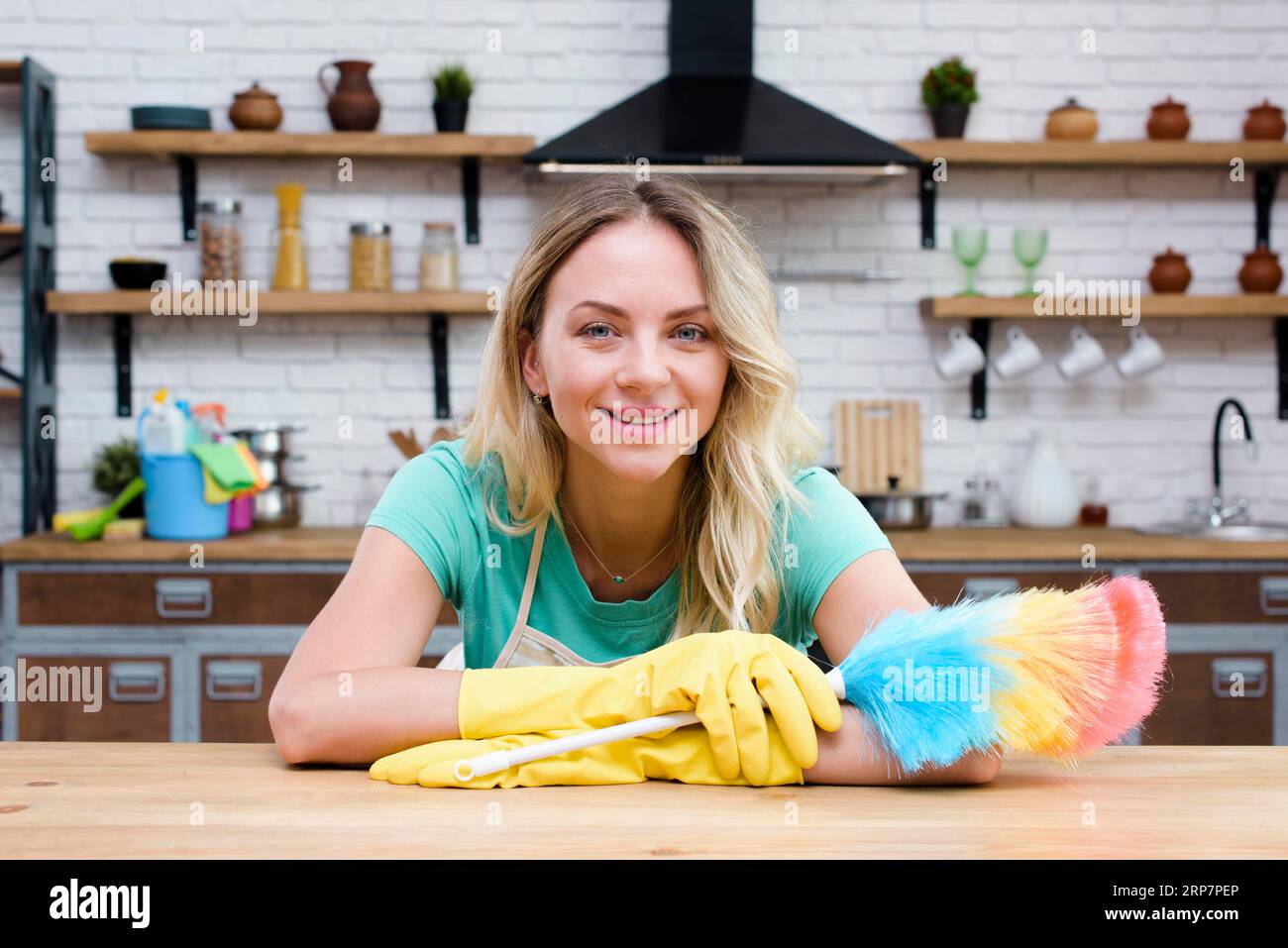 Smiling housekeeper leaning kitchen counter holding feather duster ...