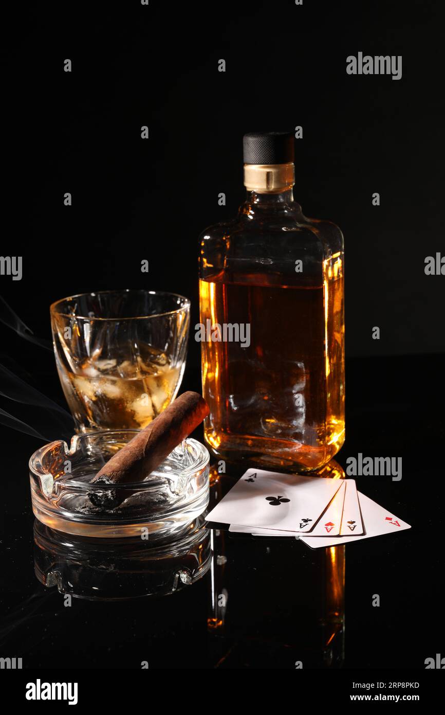 Smoldering cigar, ashtray, playing cards and whiskey on black mirror ...