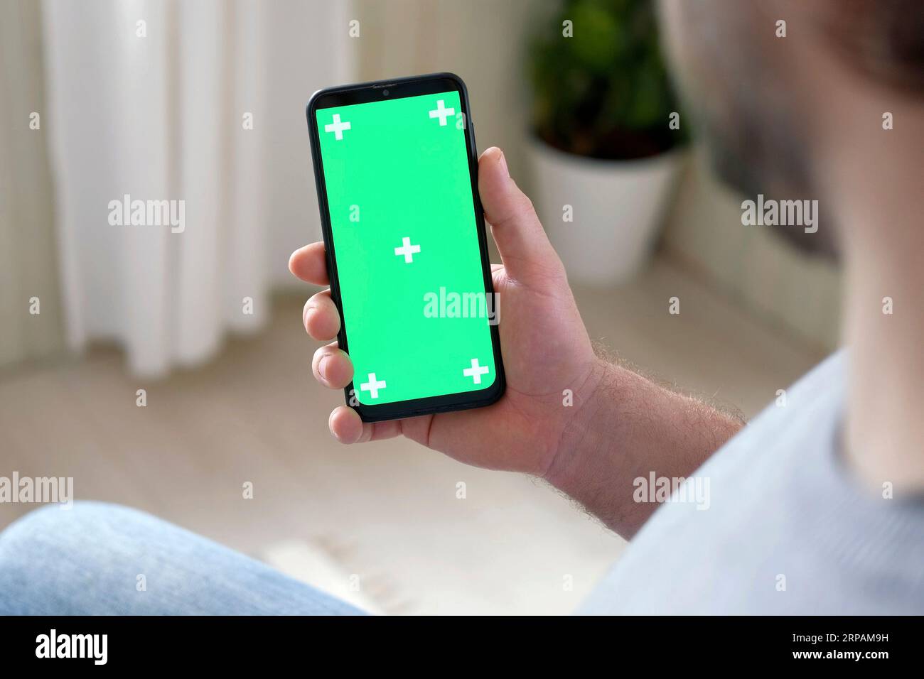 Young man sitting at home holding smartphone green mock-up screen in hand. Male person using ...