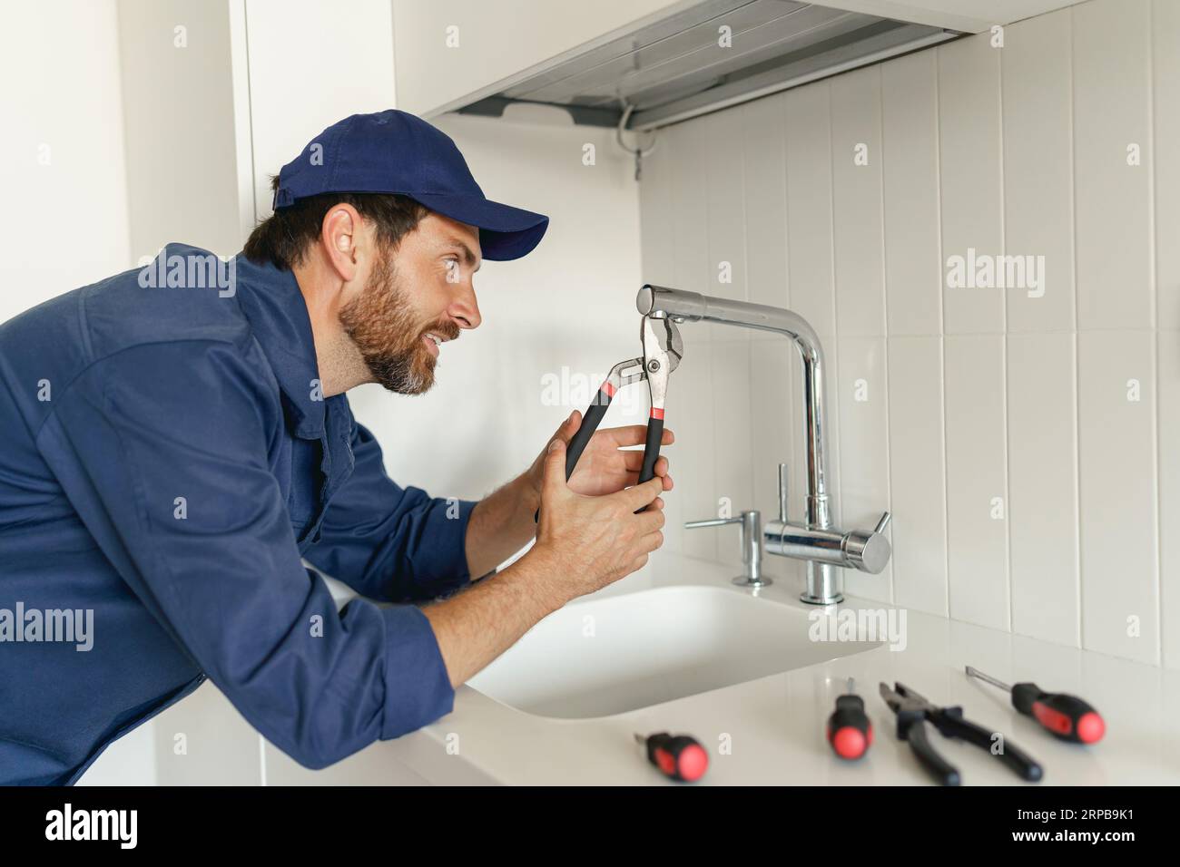 Professional plumber in blue uniform fixing broken water tap in kitchen ...