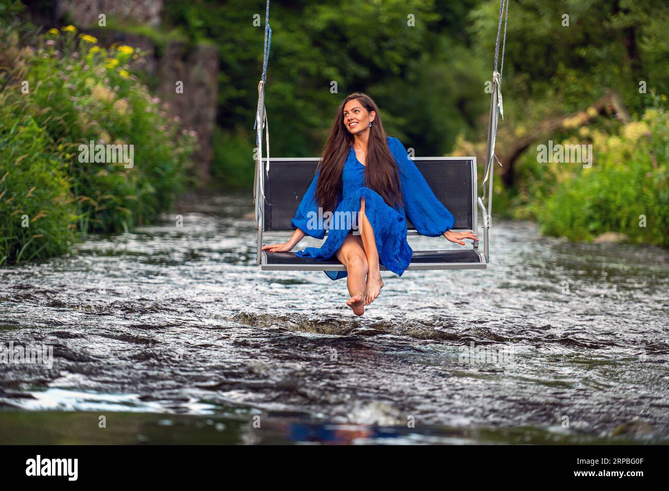a smiling young woman swings on a rope swing across a fast-flowing ...