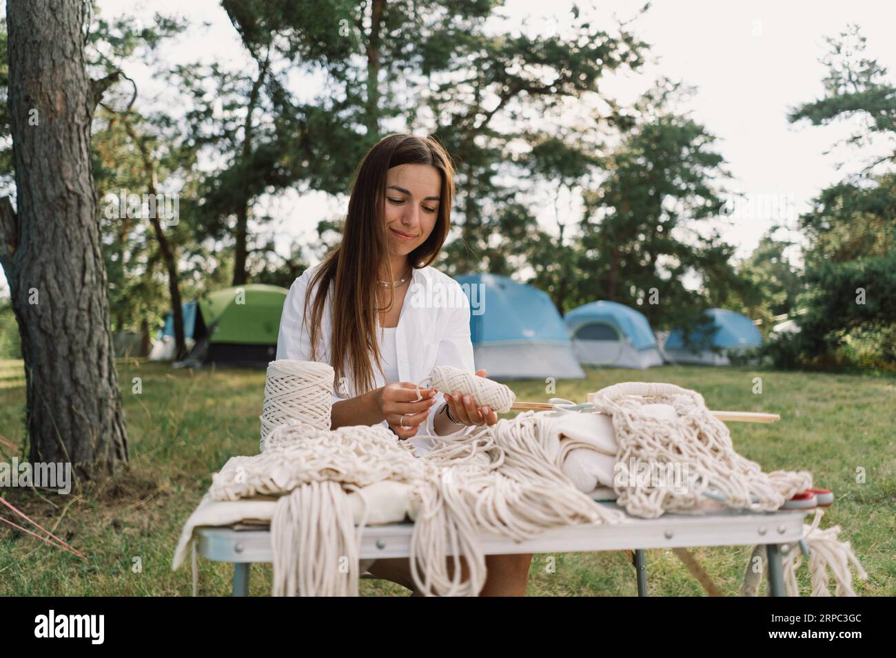 Woman knits bag using macrame technique outdoors near tents. Outdoor ...