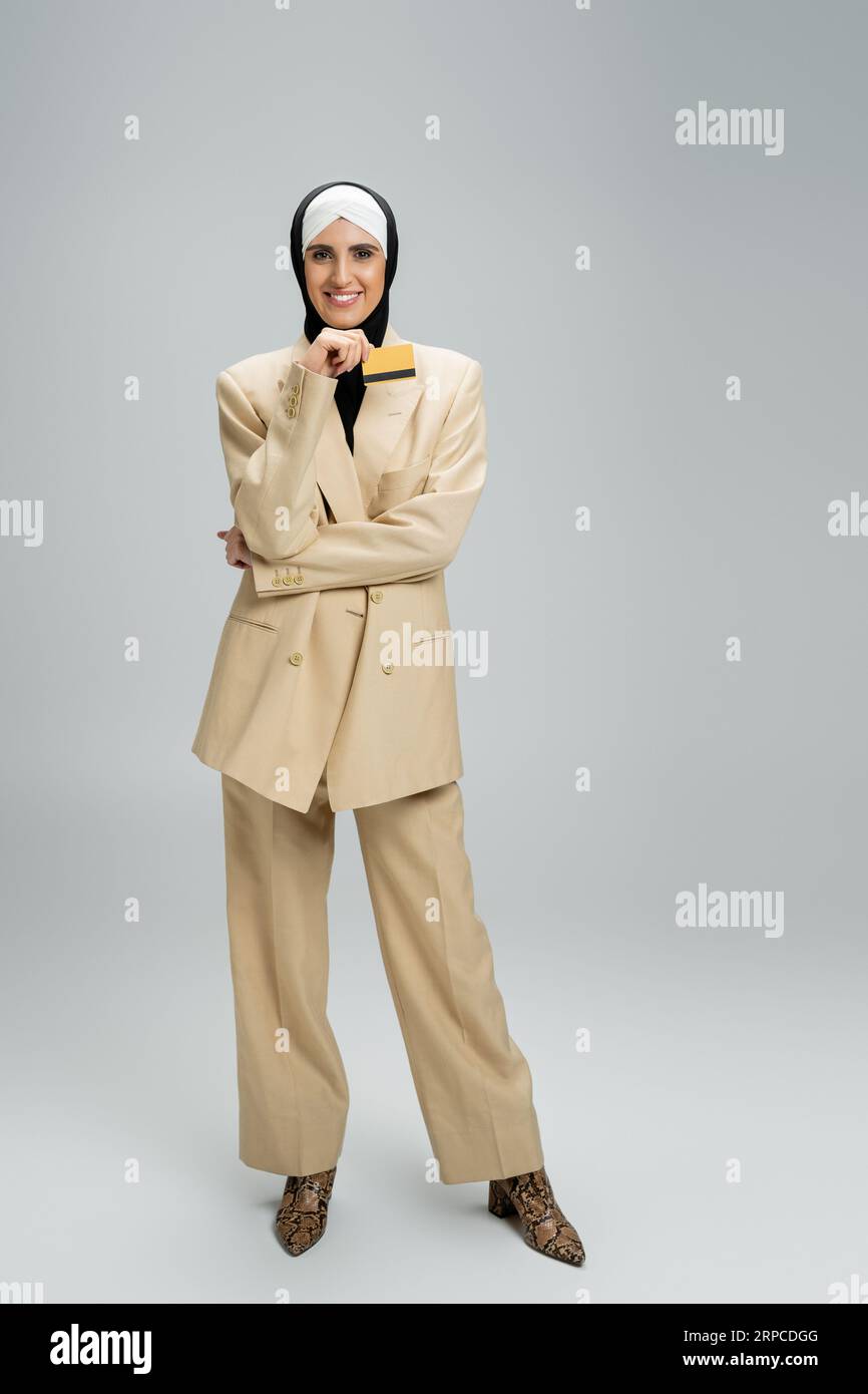positive middle eastern woman in hijab and beige suit posing with ...