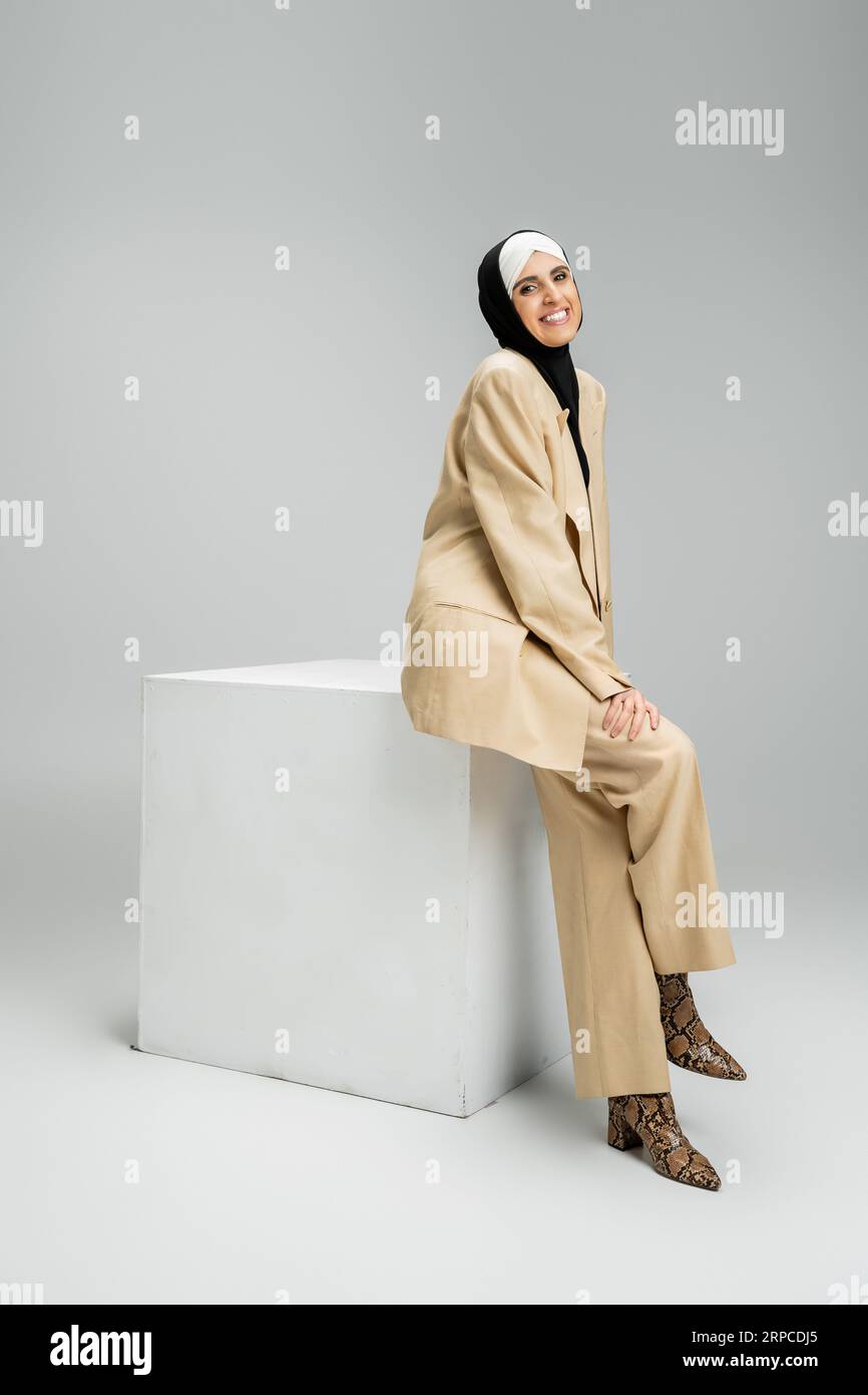 joyful muslim businesswoman in hijab and beige suit sitting on white ...