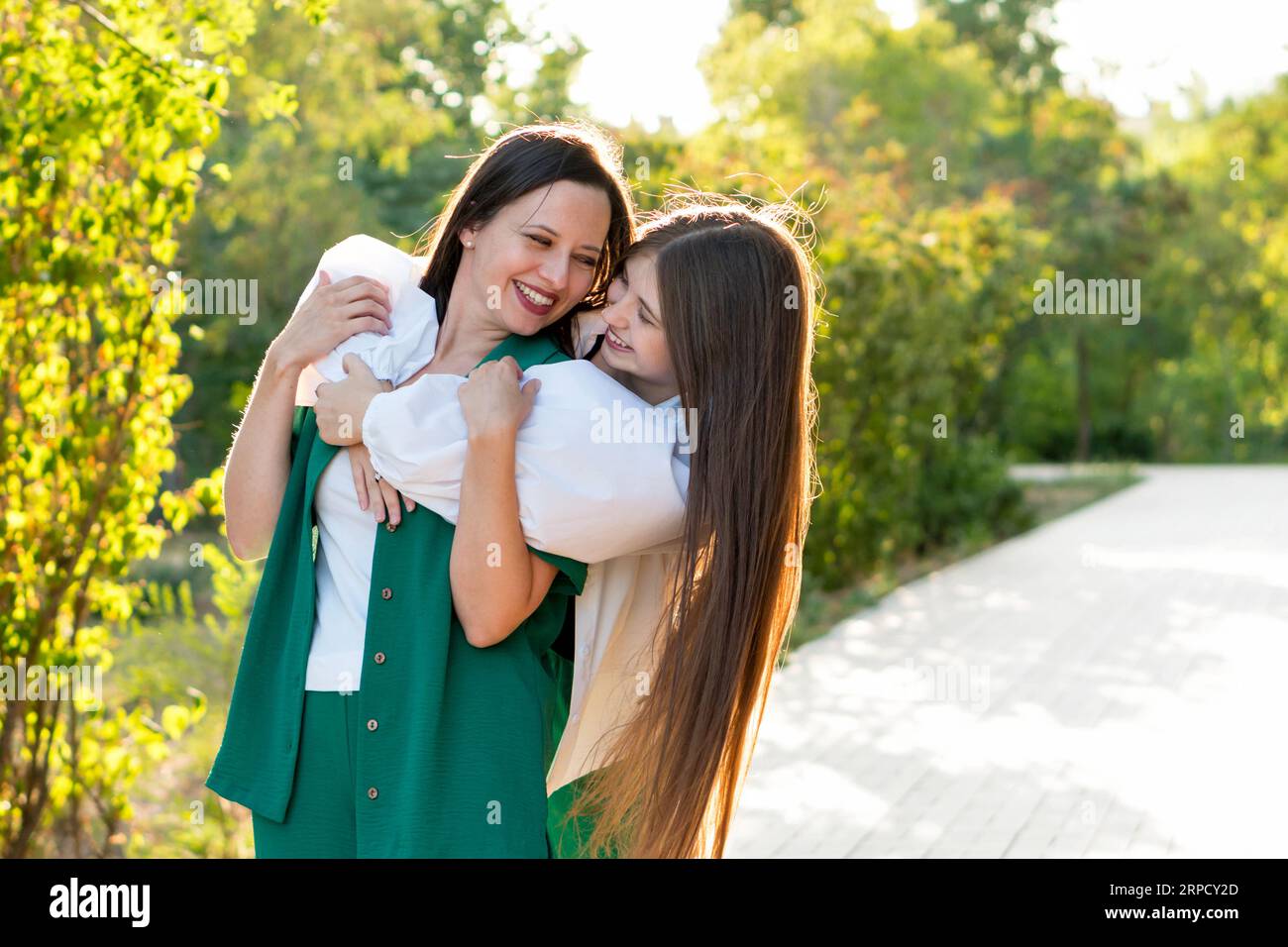 Cheerful mom and teenage daughter hugging on walk in park. Long-haired teen girl emotionally ...