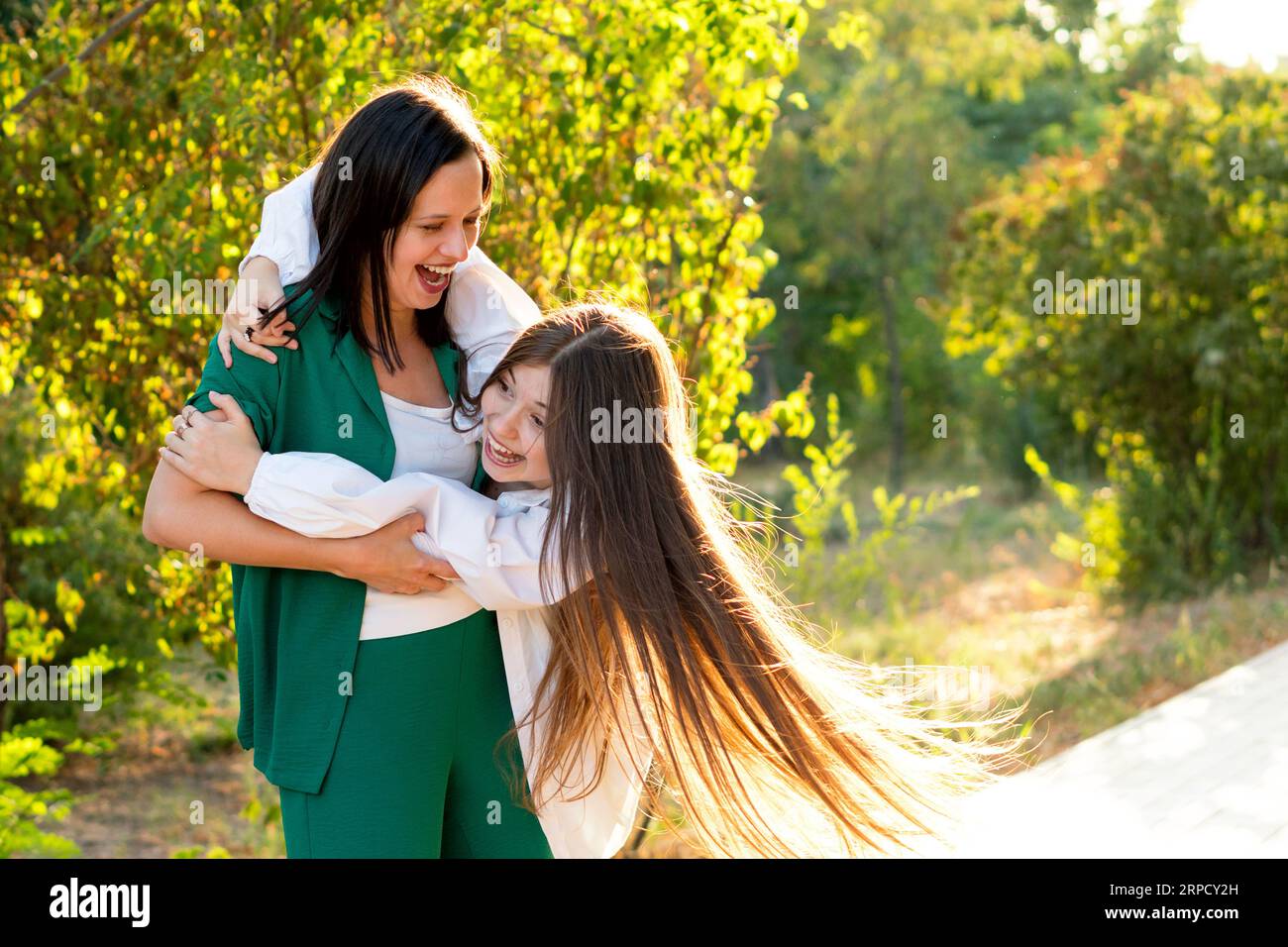 Cheerful mom and teenage daughter hugging on walk in park. Long-haired teen girl emotionally ...