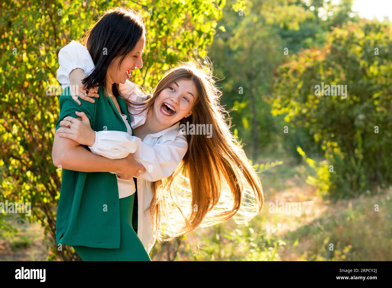 Cheerful mom and teenage daughter hugging on walk in park. Long-haired ...