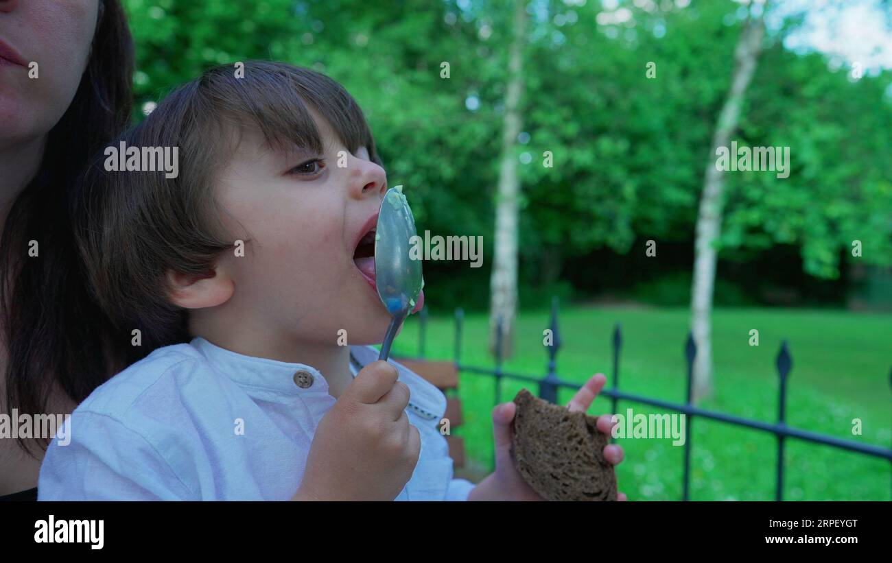 Child tasting puree food with spoon on mother's lap seated outside in ...