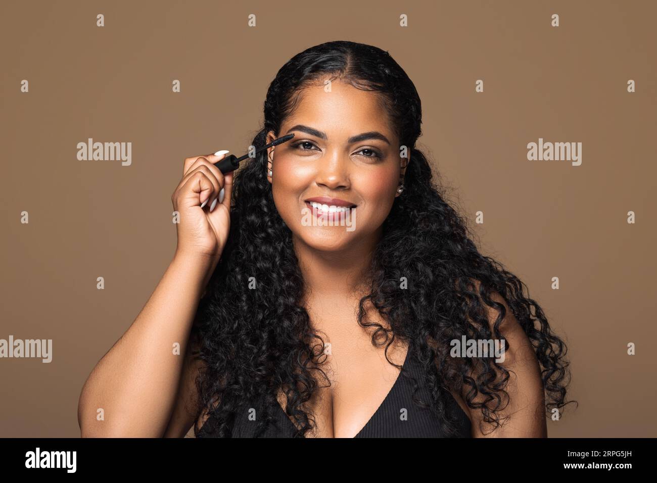 Beauty portrait of charming brazilian chubby woman applying mascara on her lashes over brown ...