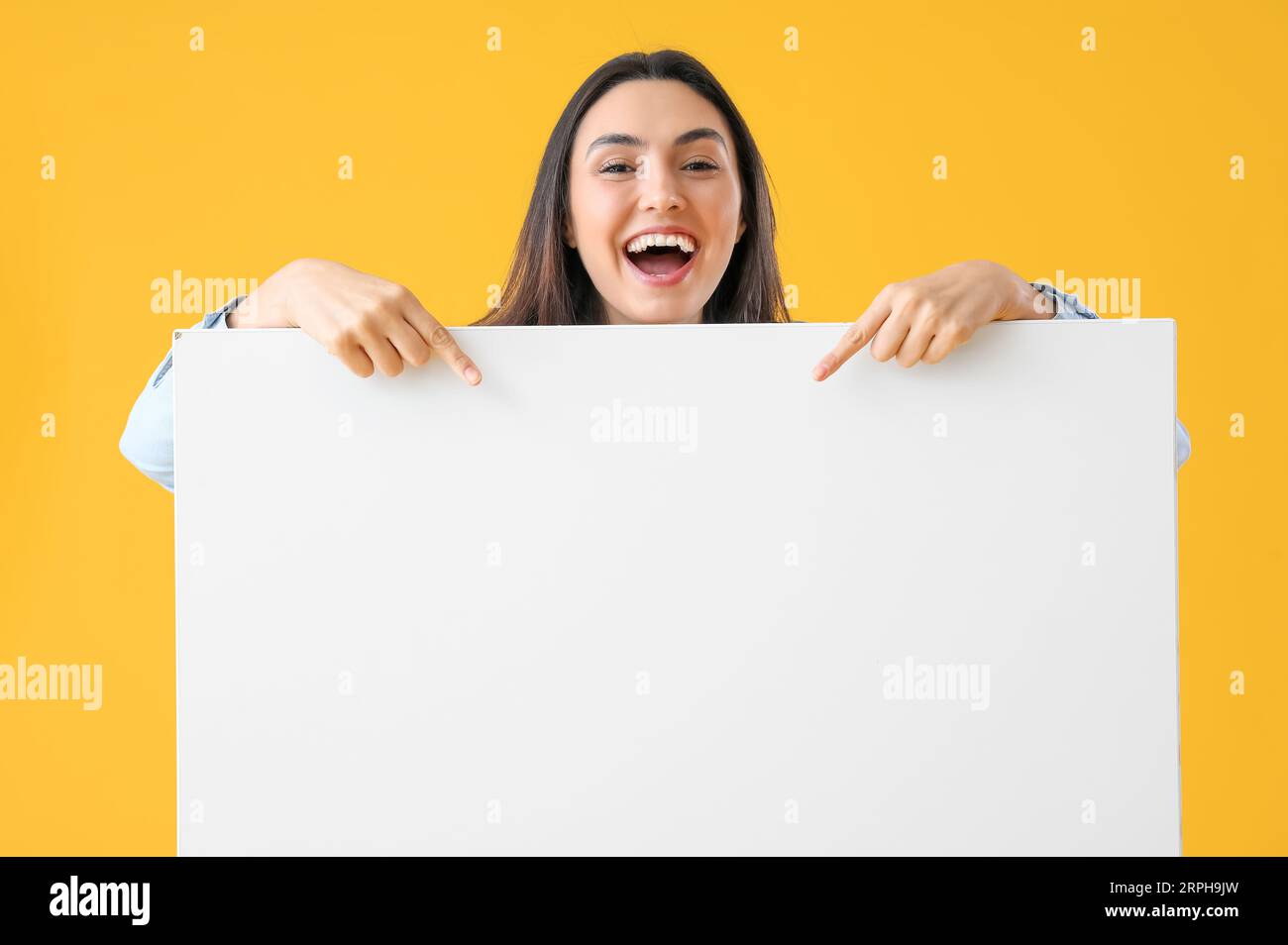 Happy young woman pointing at big blank poster on yellow background ...