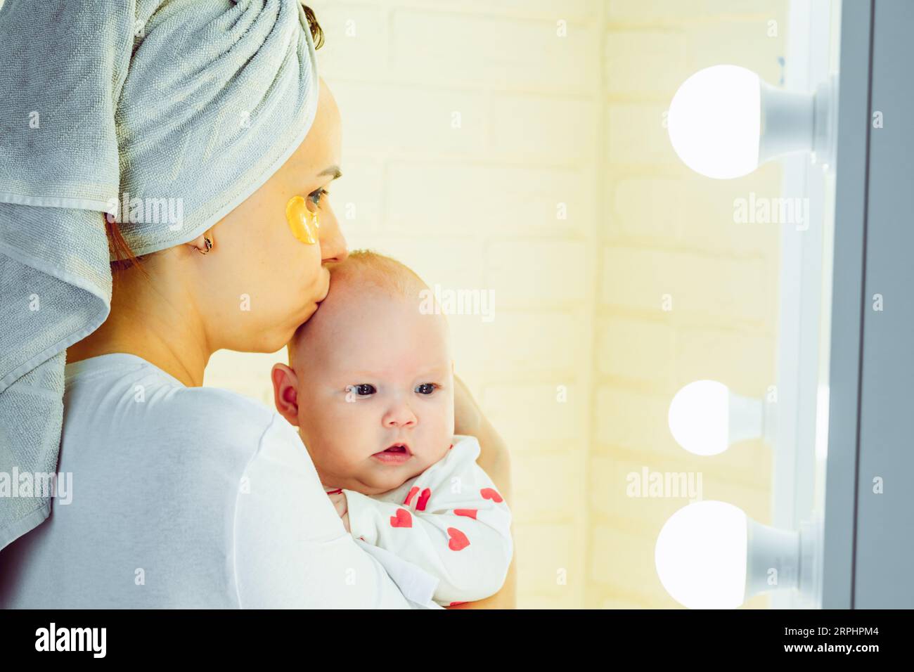 Young mother take care of her skin with under eye patches holding her newborn baby in her arms ...
