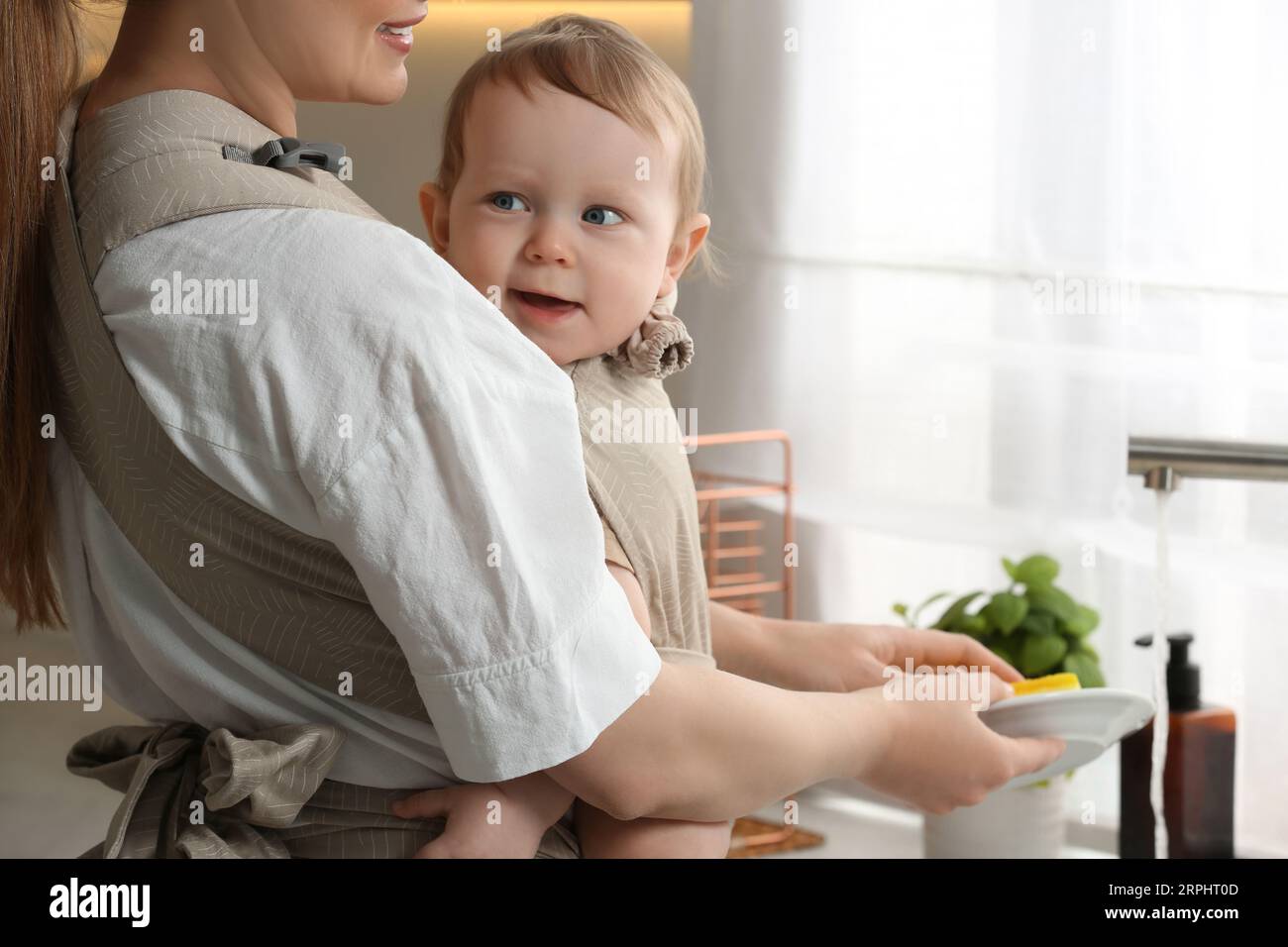 Mother holding her child in sling (baby carrier) while washing plates ...