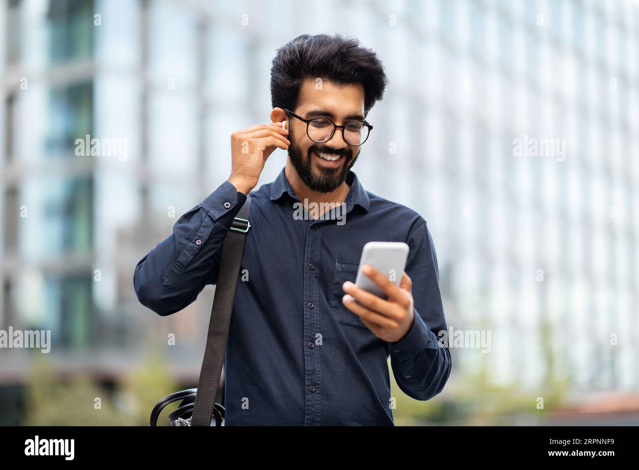 Smiling young indian man wearing earbud holding phone while walking ...