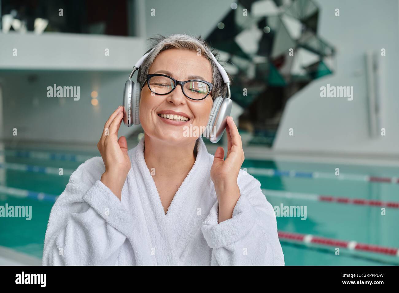 happy middle aged woman in glasses and robe listening music in wireless headphones in spa center ...