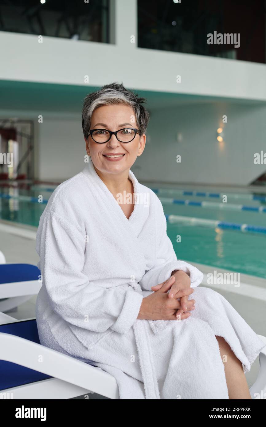 spa center concept, happy middle aged woman in glasses and robe sitting on lounger near pool ...