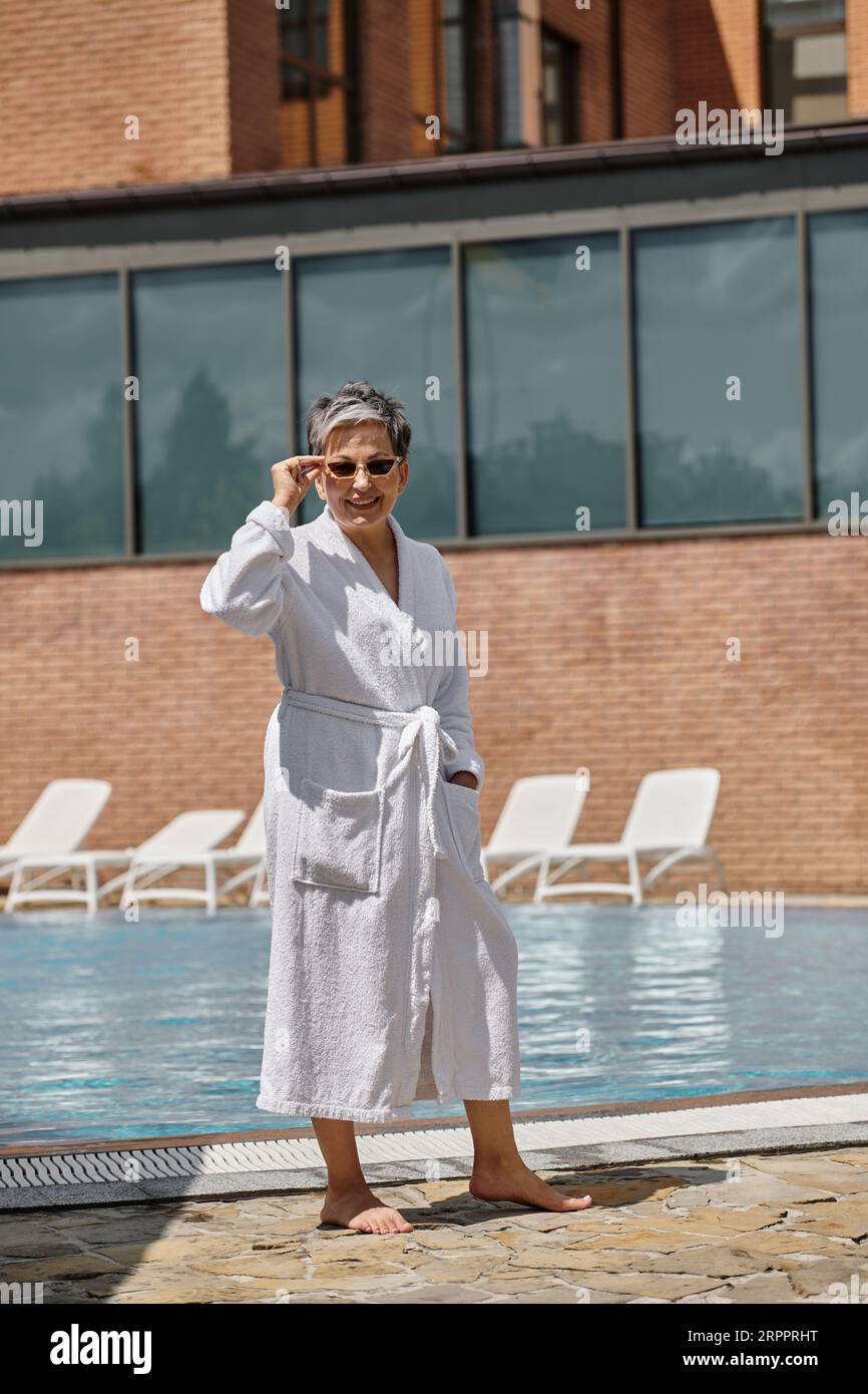 cheerful middle aged woman in white robe and sunglasses standing at ...