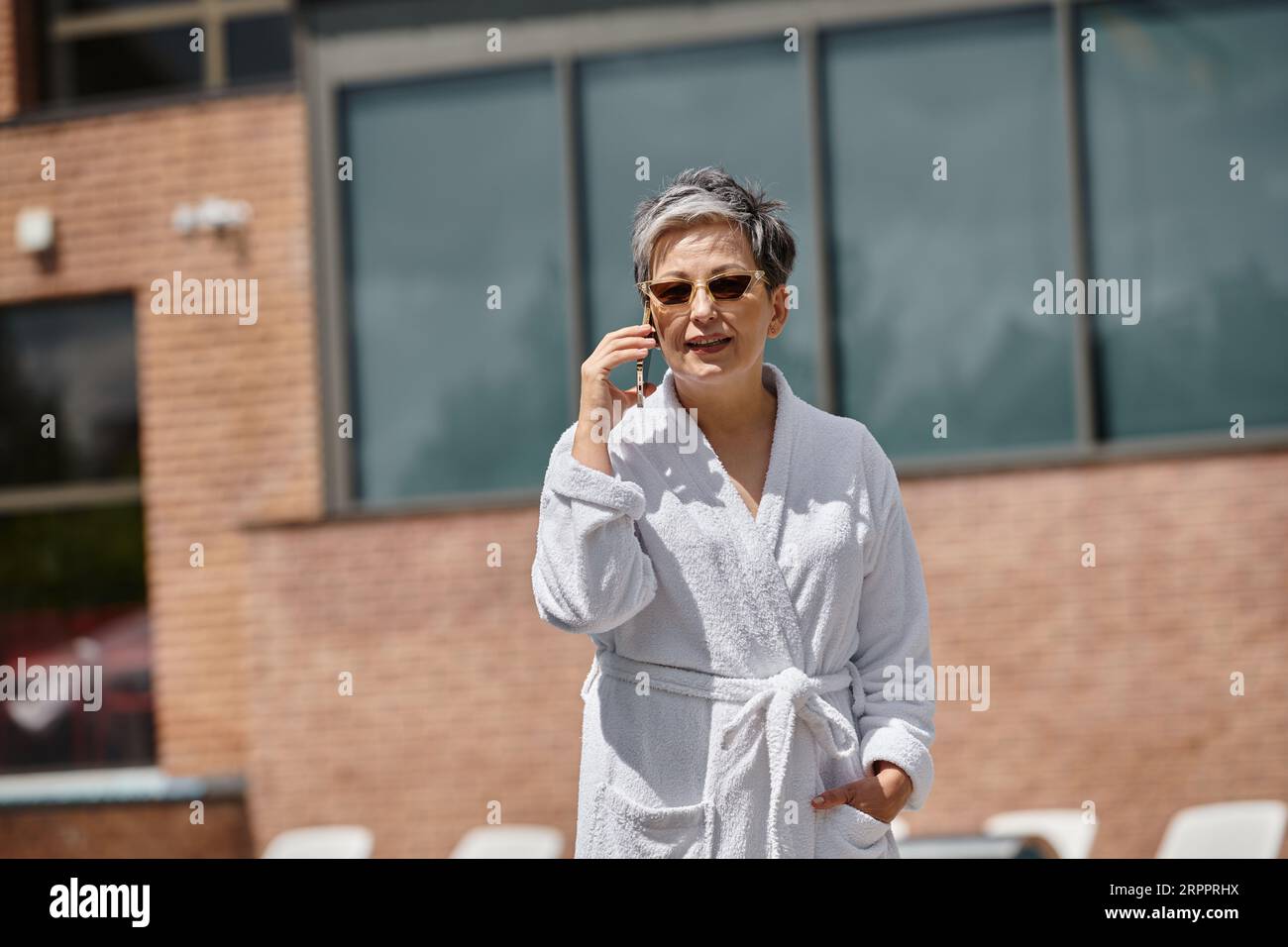 middle aged woman in white robe and sunglasses talking on smartphone at poolside in luxury ...