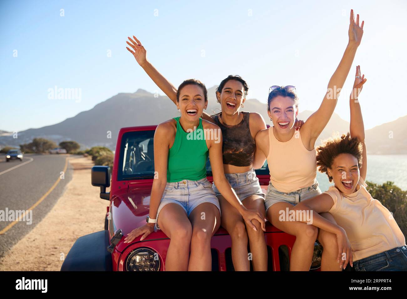 Portrait Of Laughing Female Friends Sitting On Hood Of Open Top Car On ...